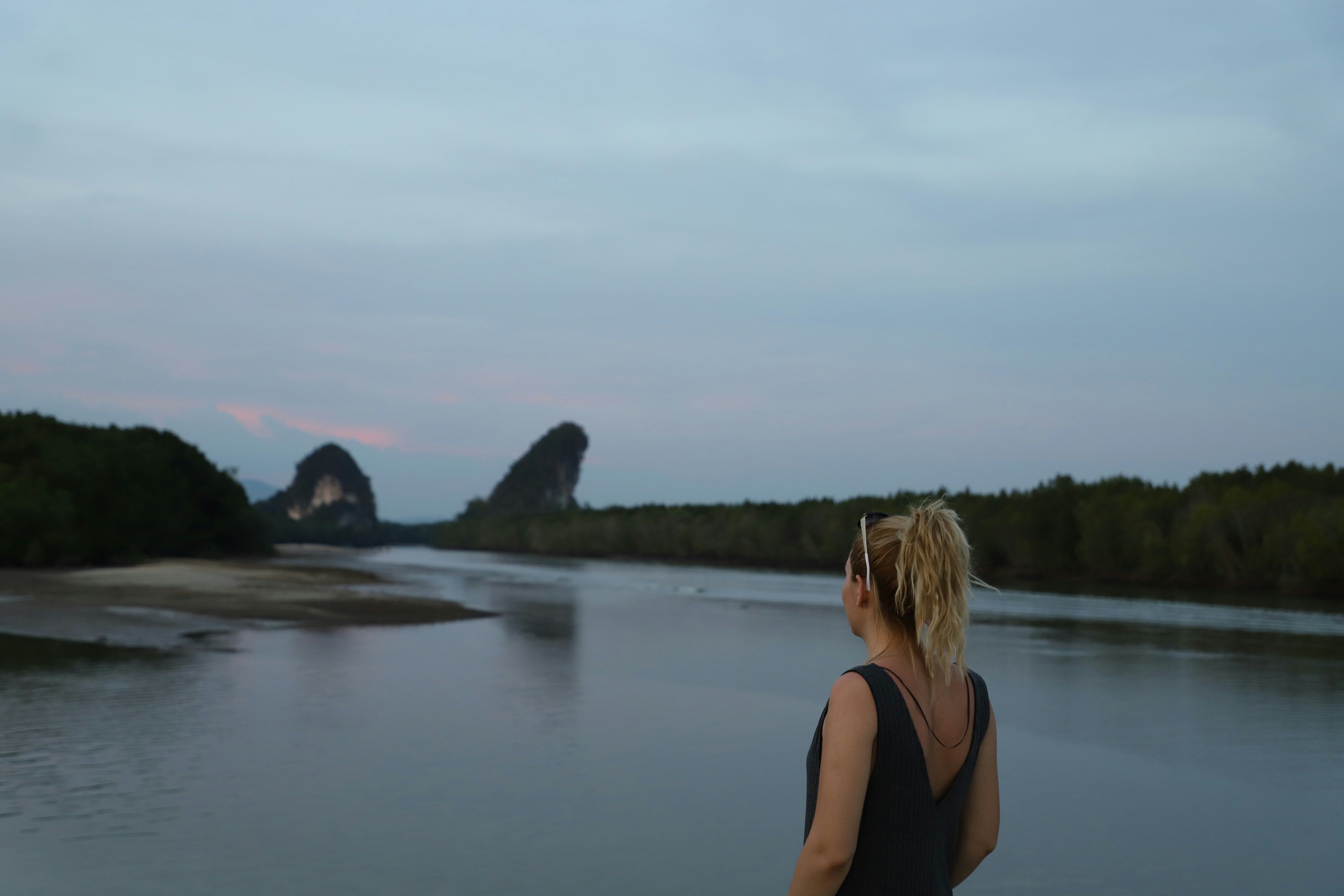 Person gazing across a serene river toward distant rocky formations under a twilight sky.