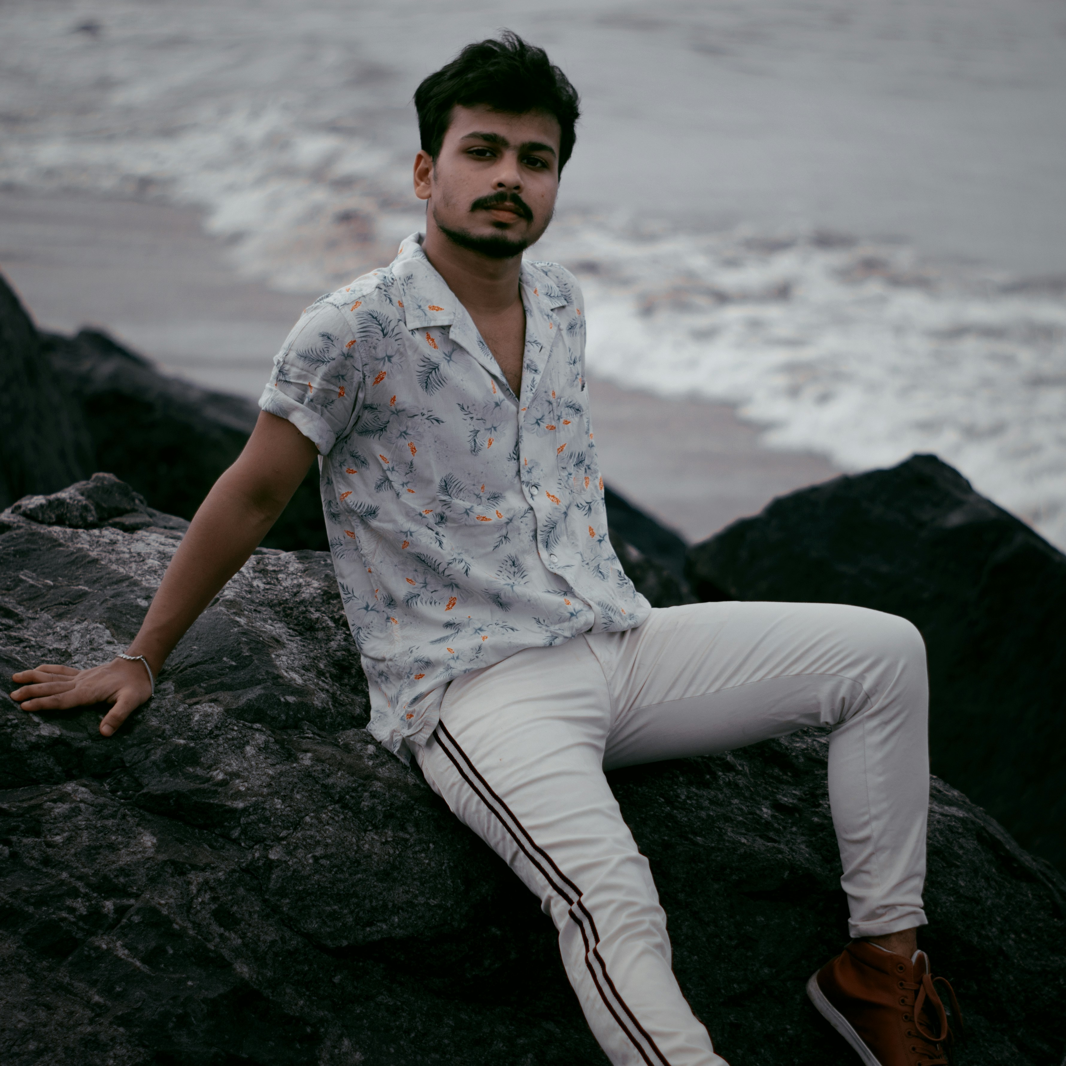 A young man in a patterned shirt sits on a rocky beach, gazing thoughtfully at the waves lapping at the shore.