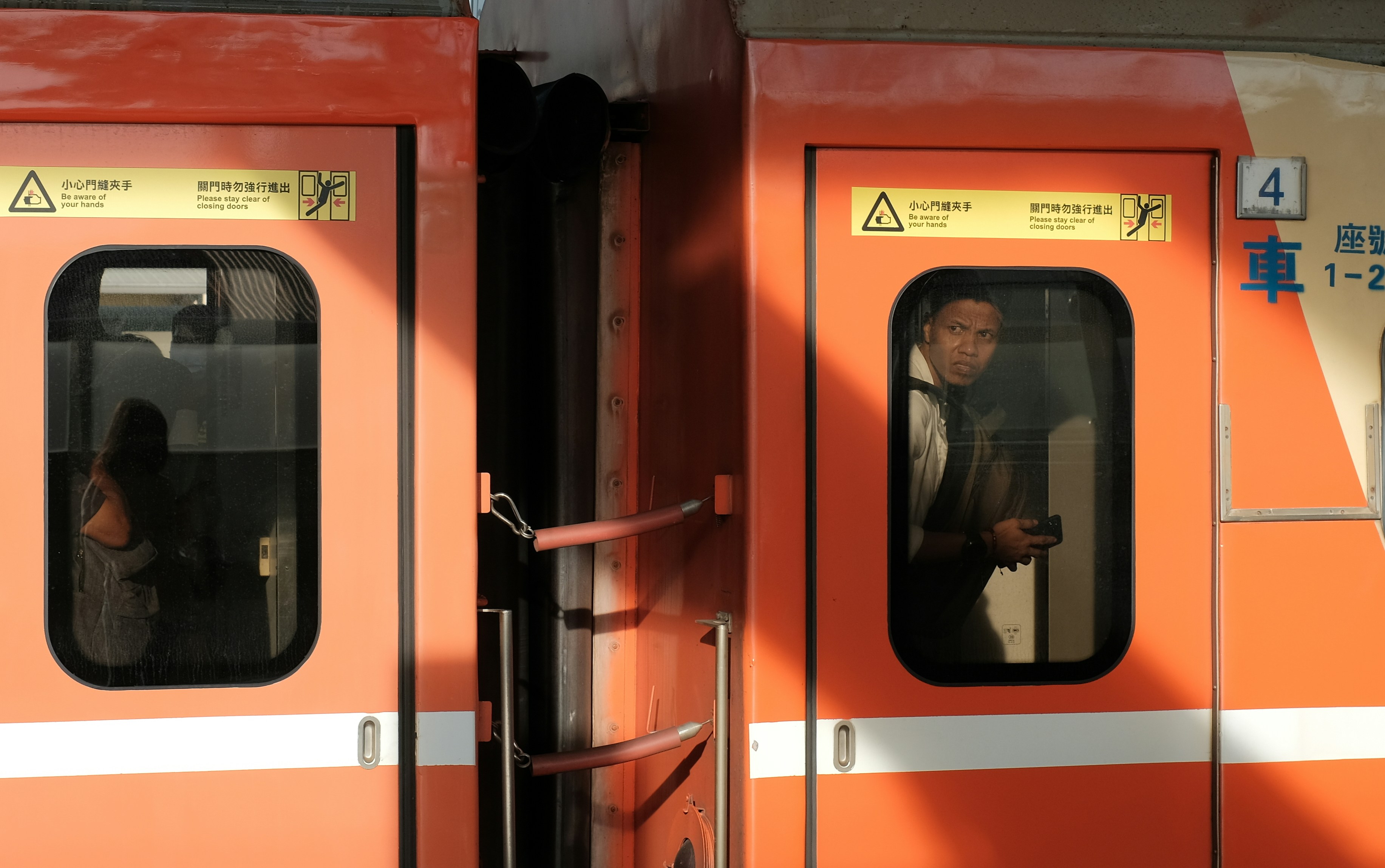A commuter gazes thoughtfully from a subway train window, framed by vibrant orange doors. The reflection of another passenger adds depth to the scene.