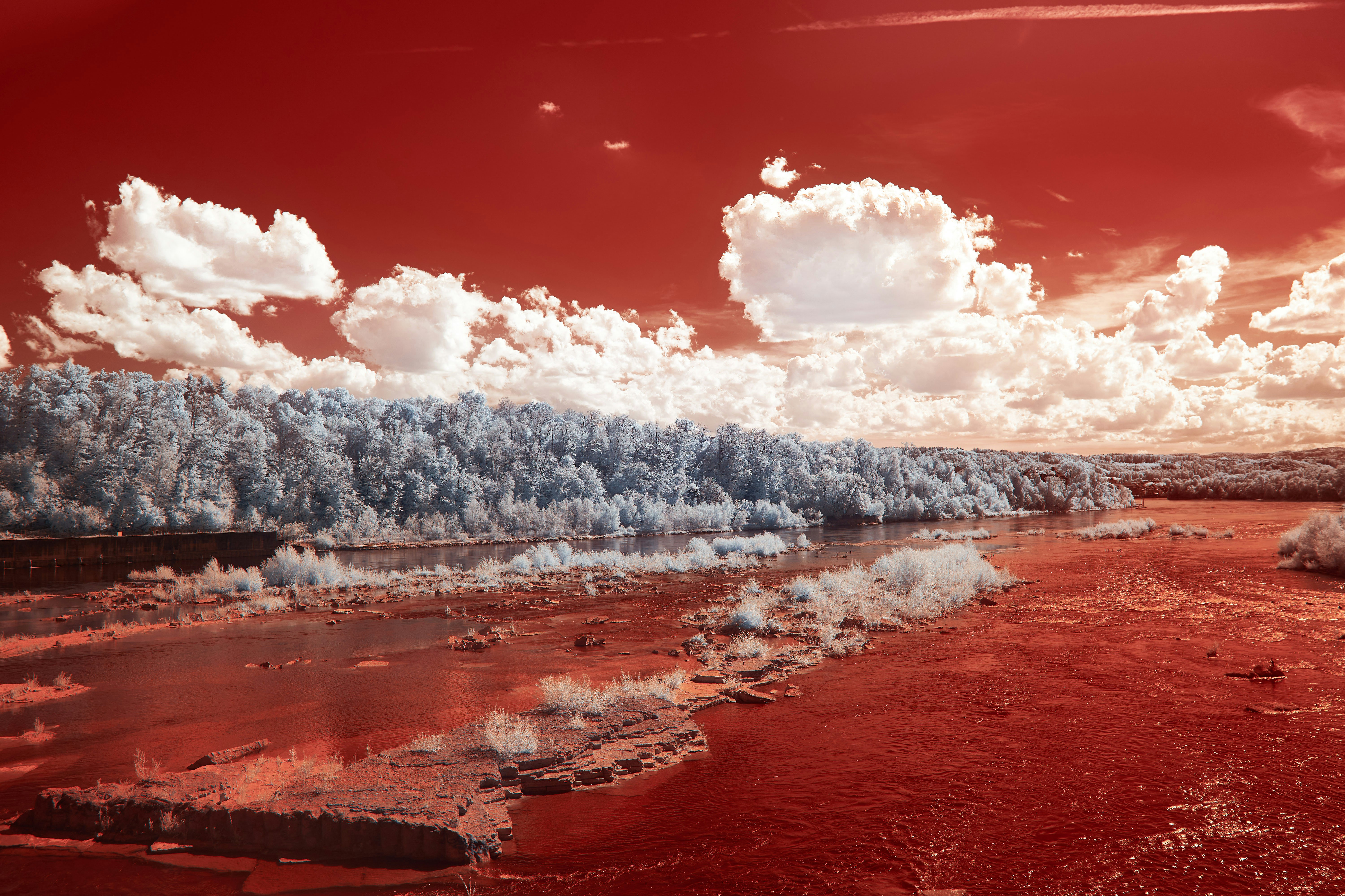 Infrared landscape with vibrant red skies and river reflecting lush trees and clouds.
