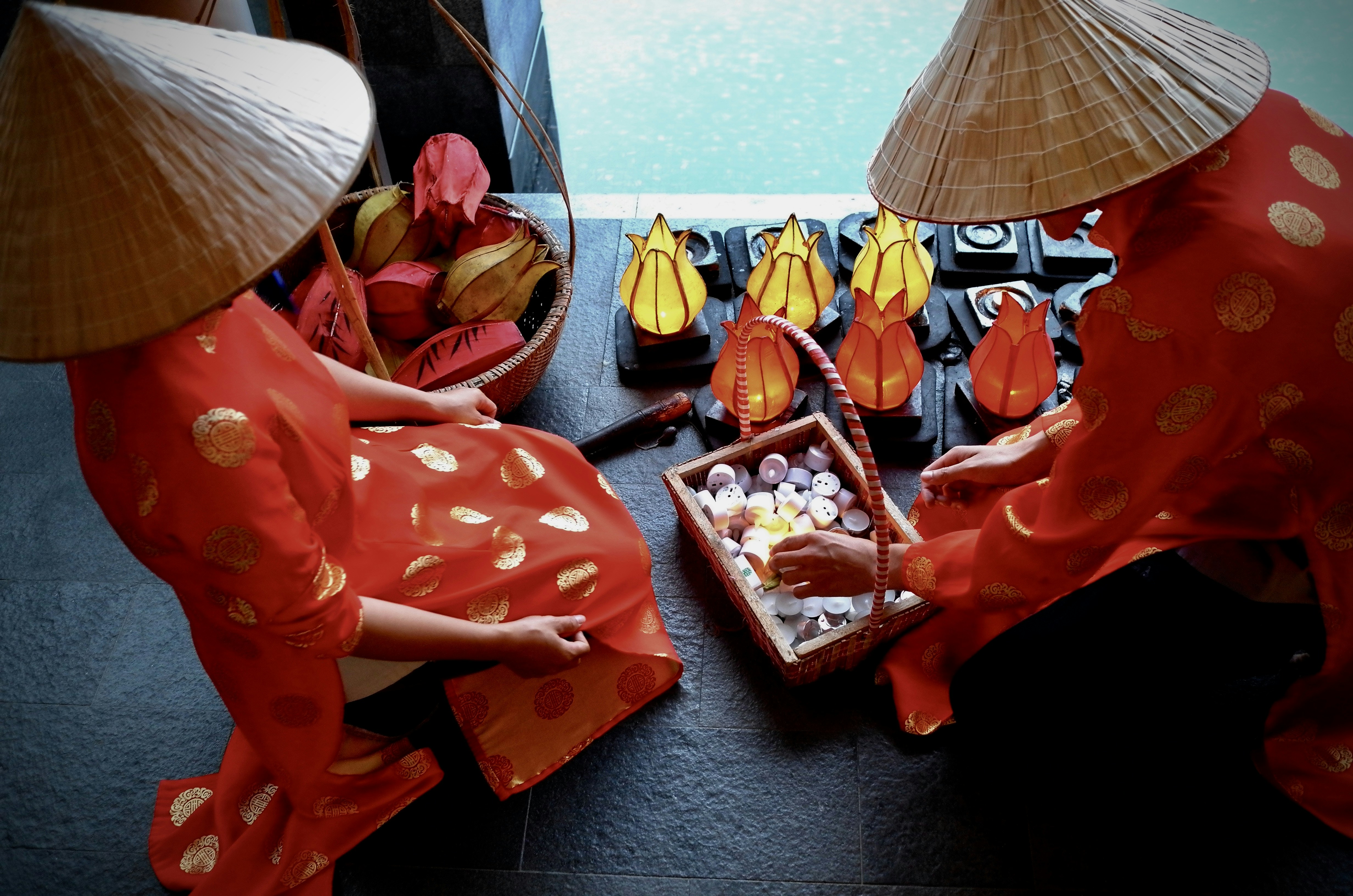 Two figures in red áo dài with conical hats sit on a dark surface, sharing a box of small fruits as lanterns float nearby by a seaside background.