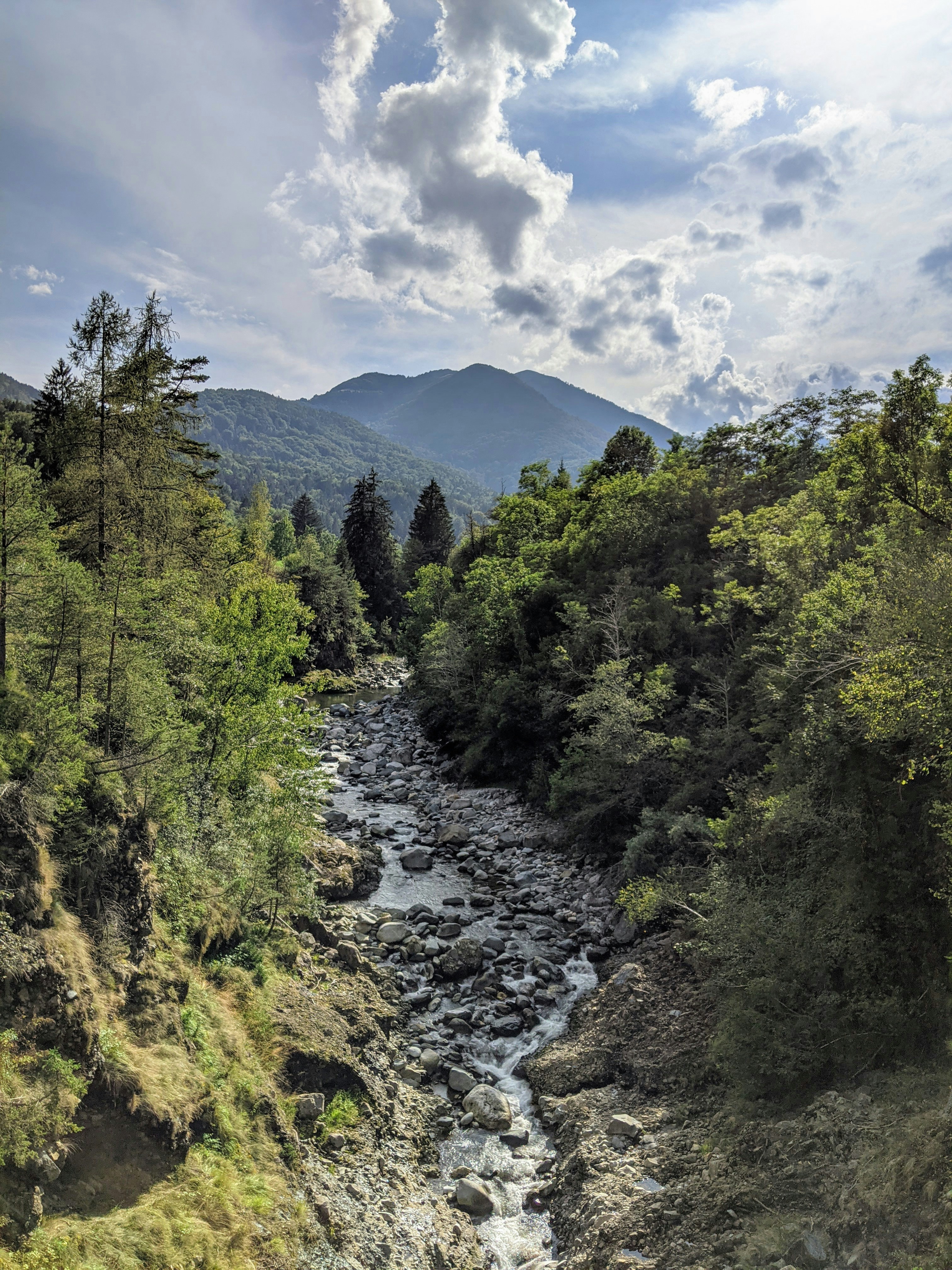 A winding stream flows through a lush landscape surrounded by mountains and trees under a cloudy sky.