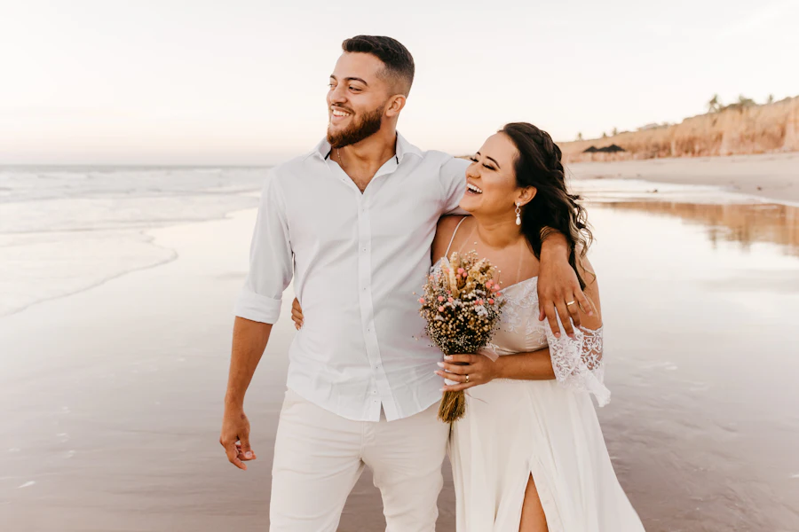 A smiling bride and groom walk arm in arm along a calm beach, with the bride holding a bouquet and seaside cliffs glowing in the background.