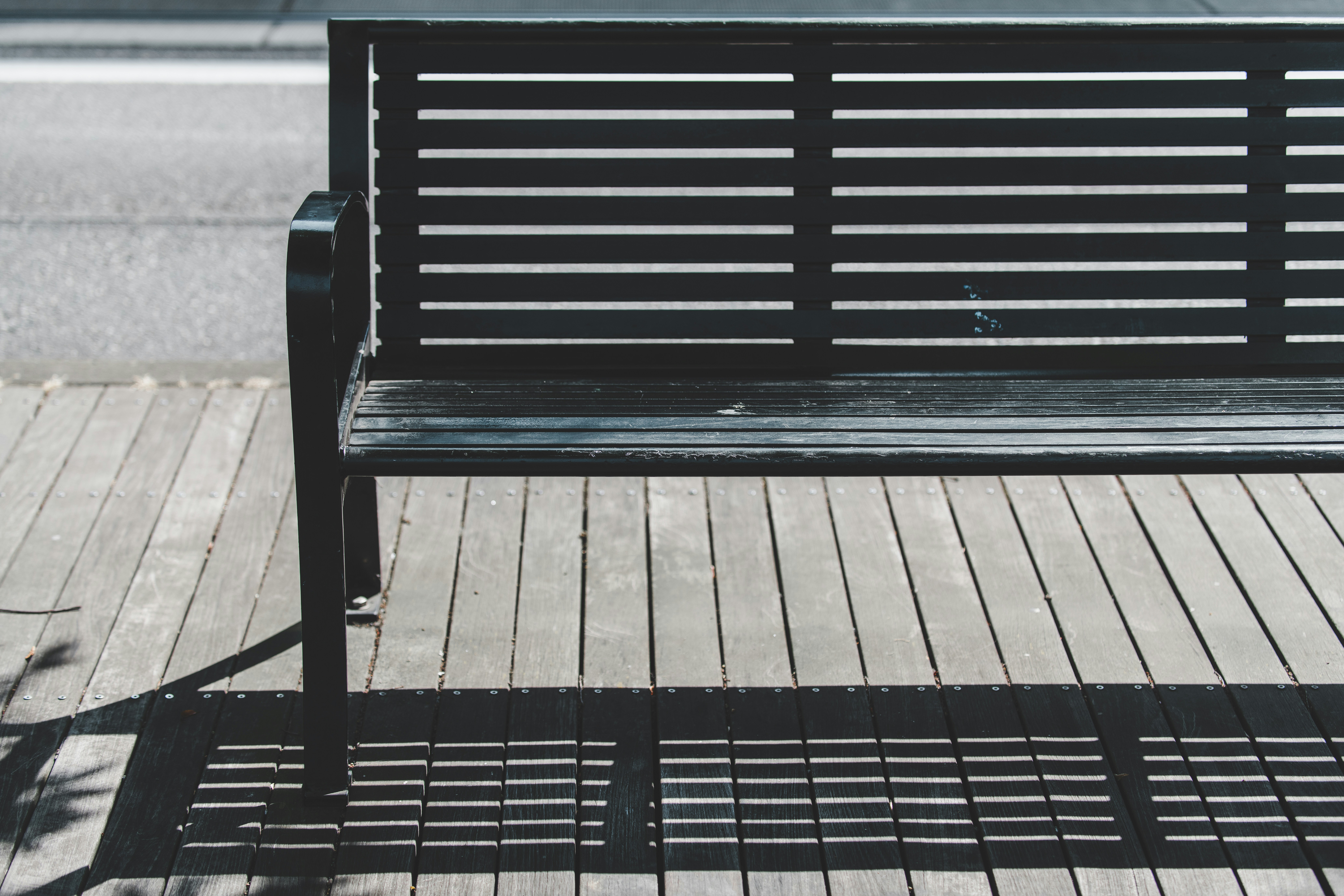 A minimalist black bench casting intricate shadows on a wooden deck, illuminated by soft sunlight. The scene evokes a sense of tranquility and solitude.