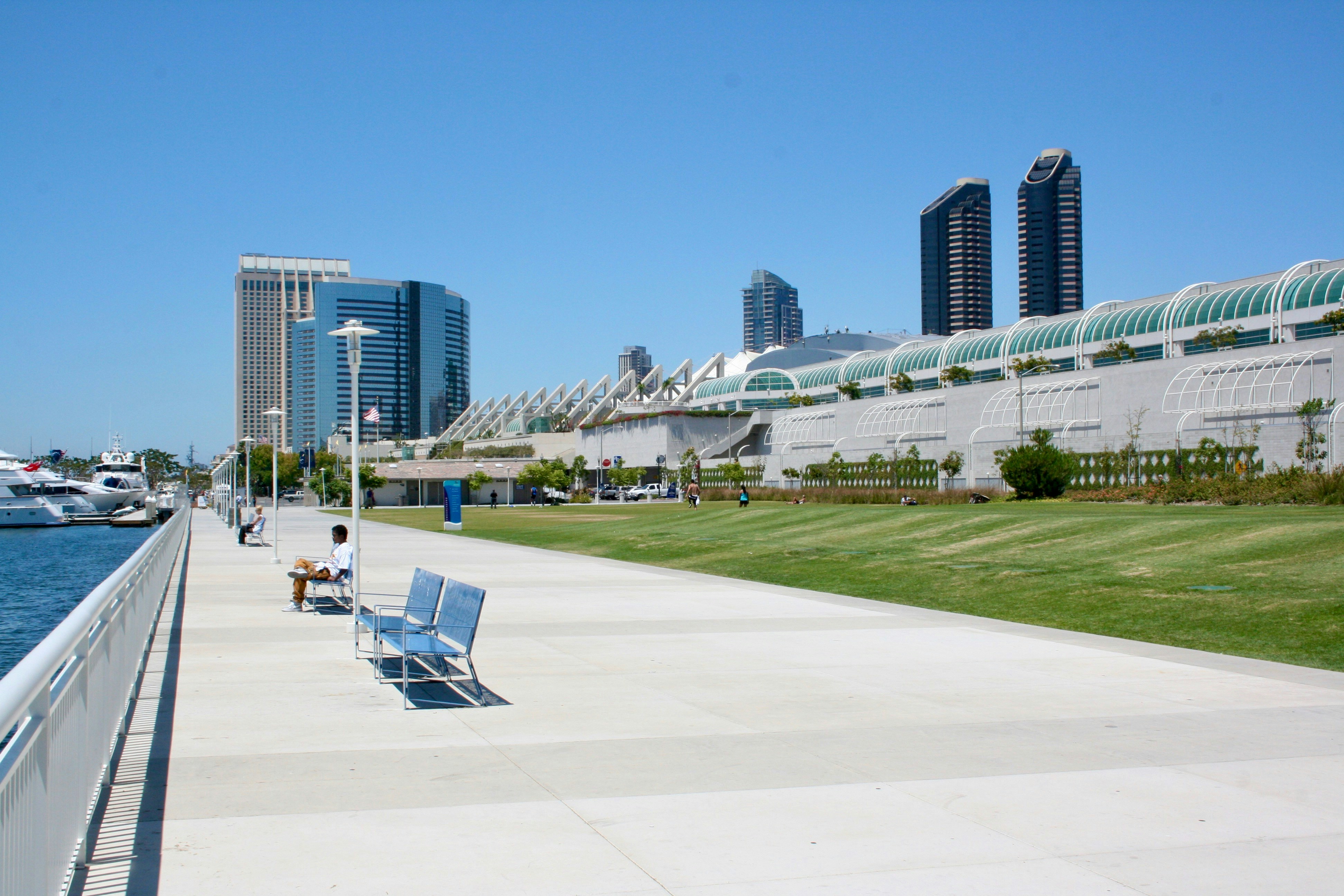 Blue metal benches line a waterfront promenade near city buildings under a clear sky.
