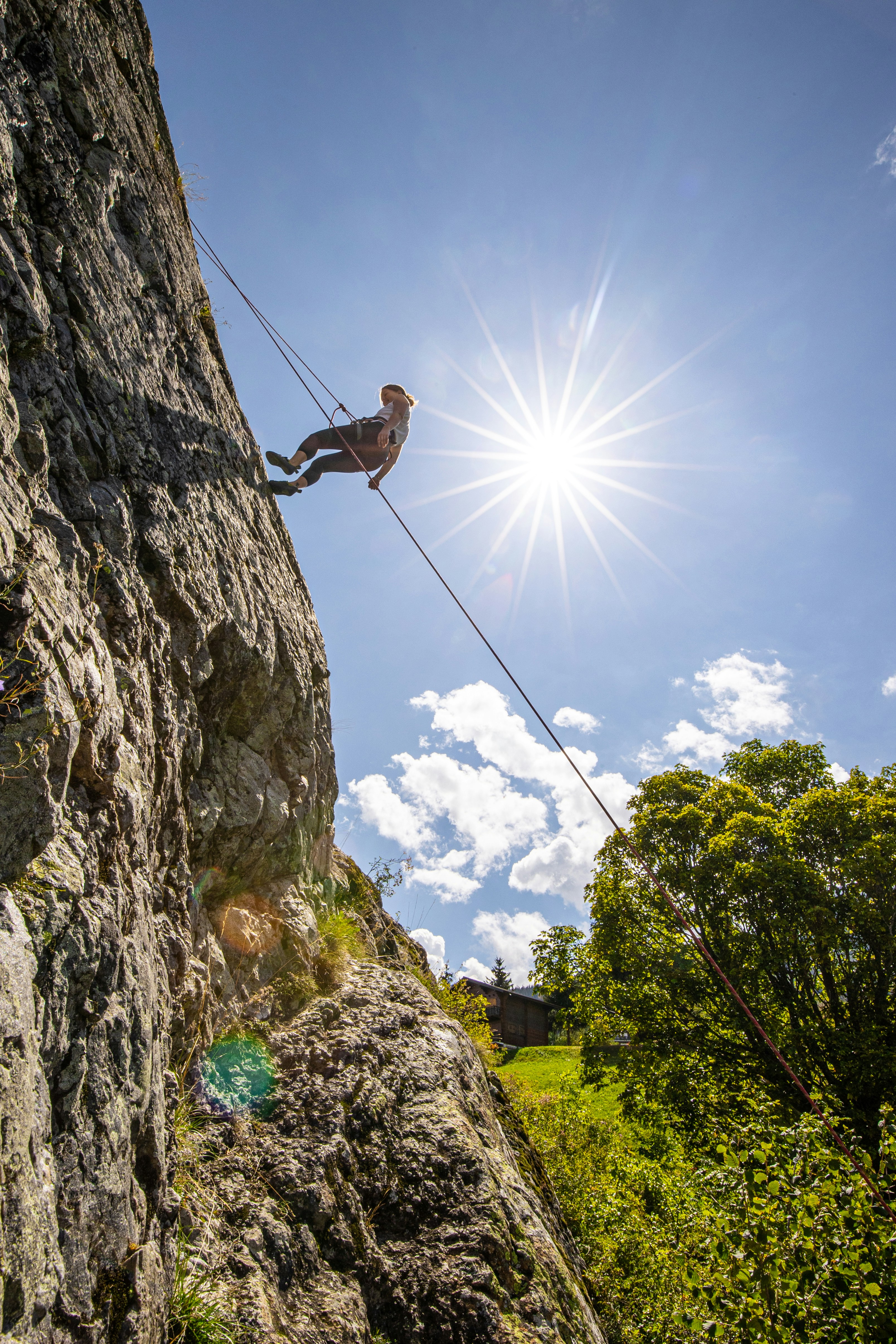 A climber skillfully rappelling down a rocky cliff under a radiant sun, with fluffy clouds scattered in the blue sky.