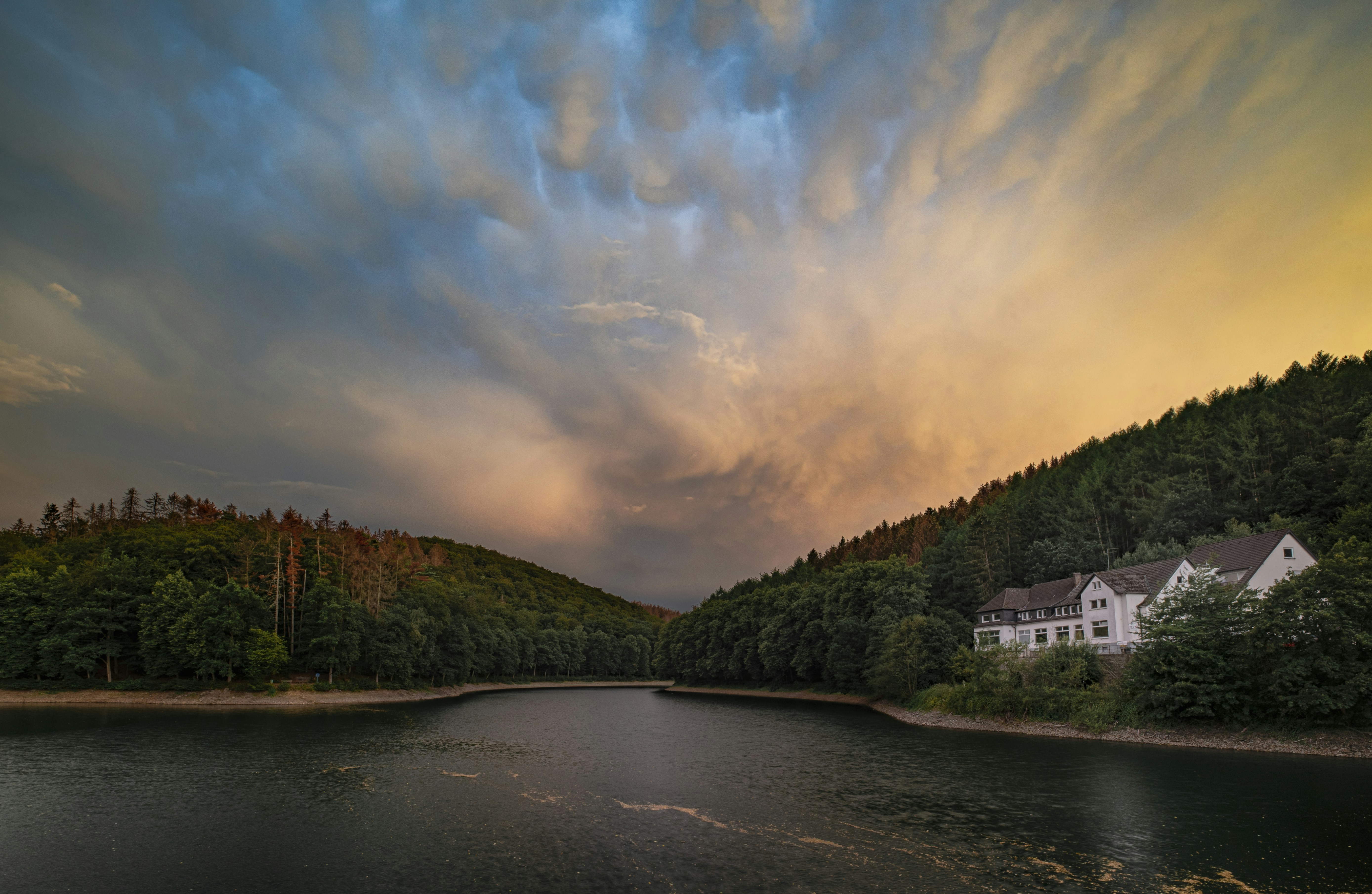 Expansive sky with dynamic clouds over a tranquil river flanked by lush forests and a distant house at sunset.
