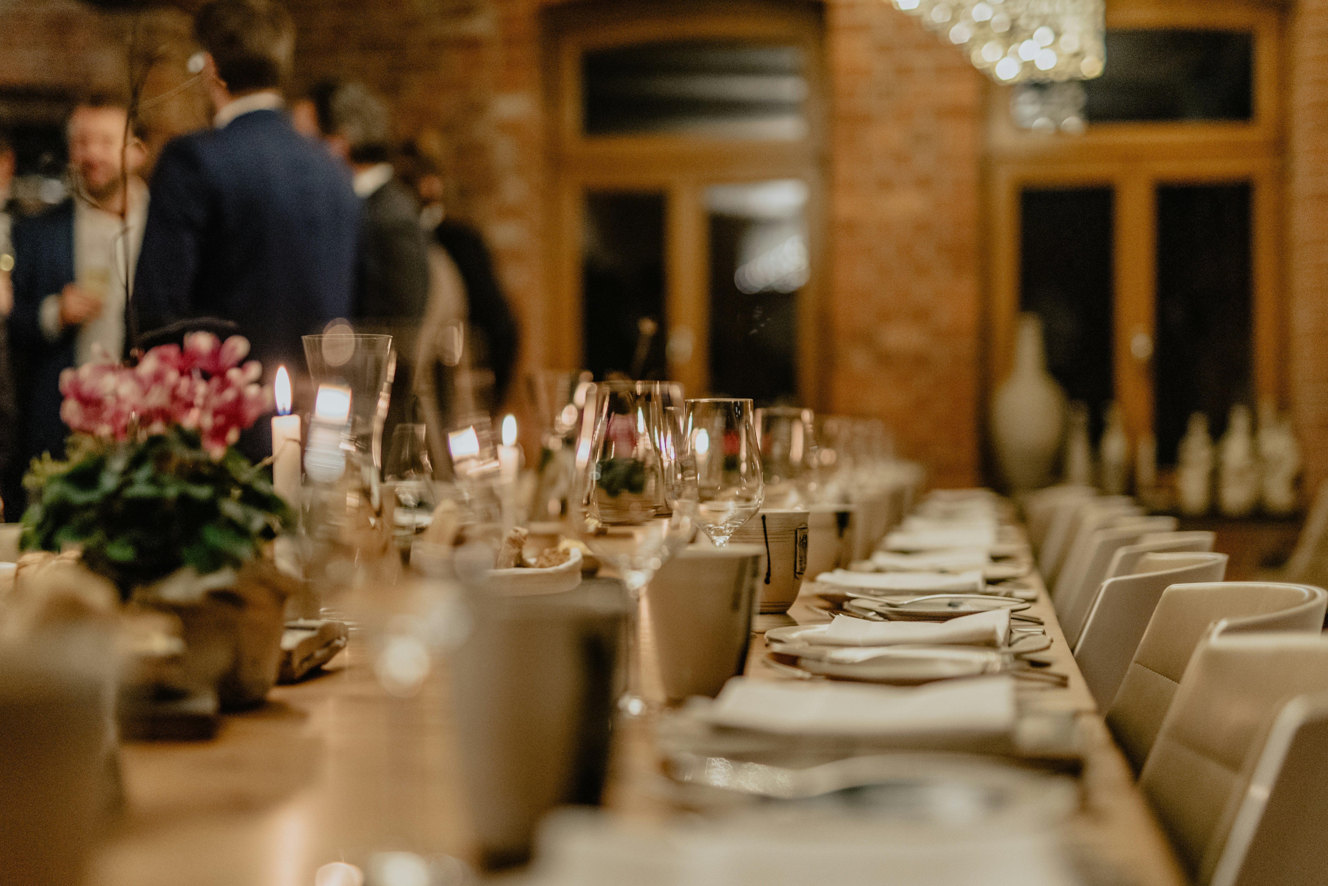 Long dining table elegantly set with glassware and floral arrangements in a warmly lit room with exposed brick walls.