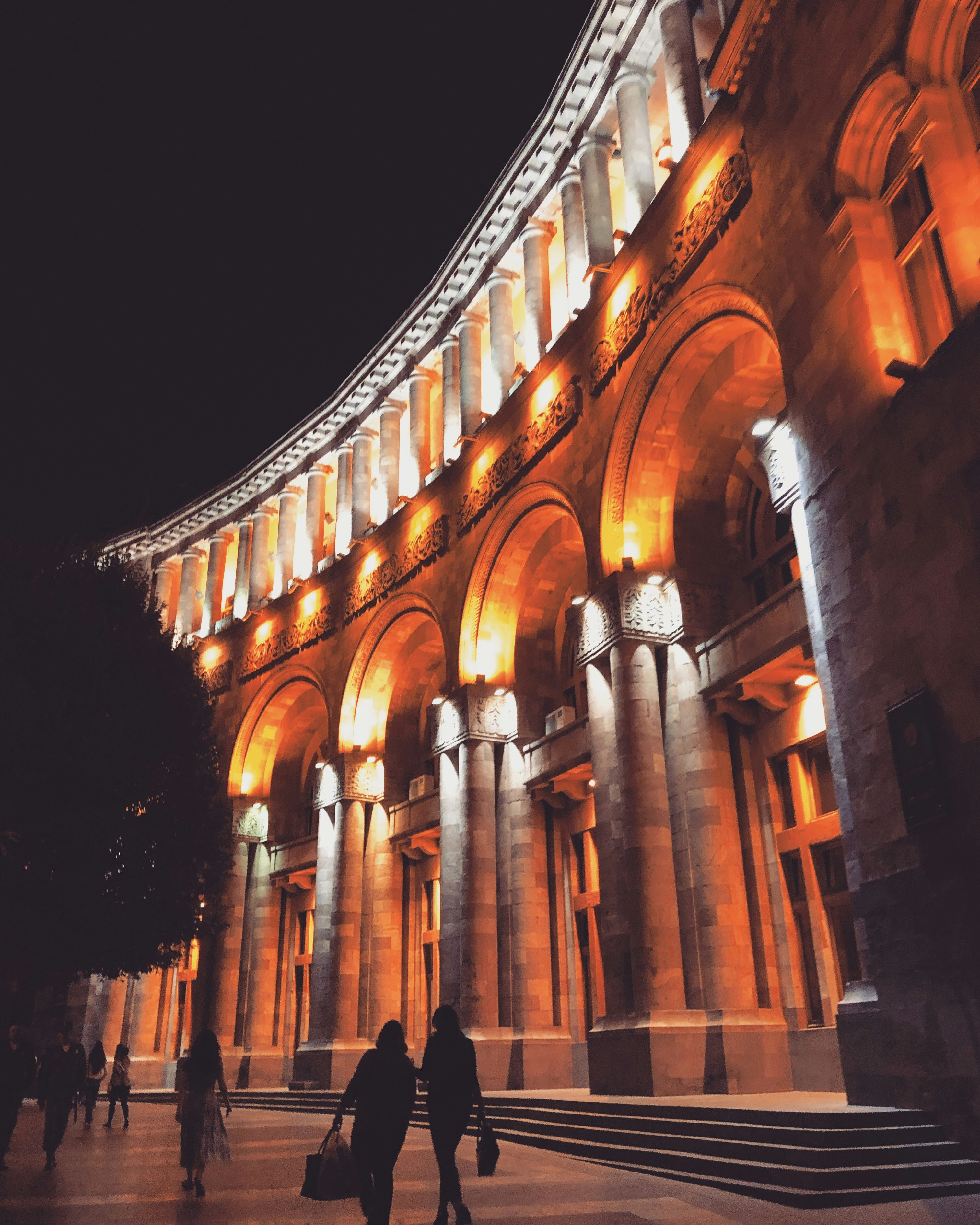 Elegant arched columns glow warmly under night lights, framing a serene walkway where silhouettes of people pass by.