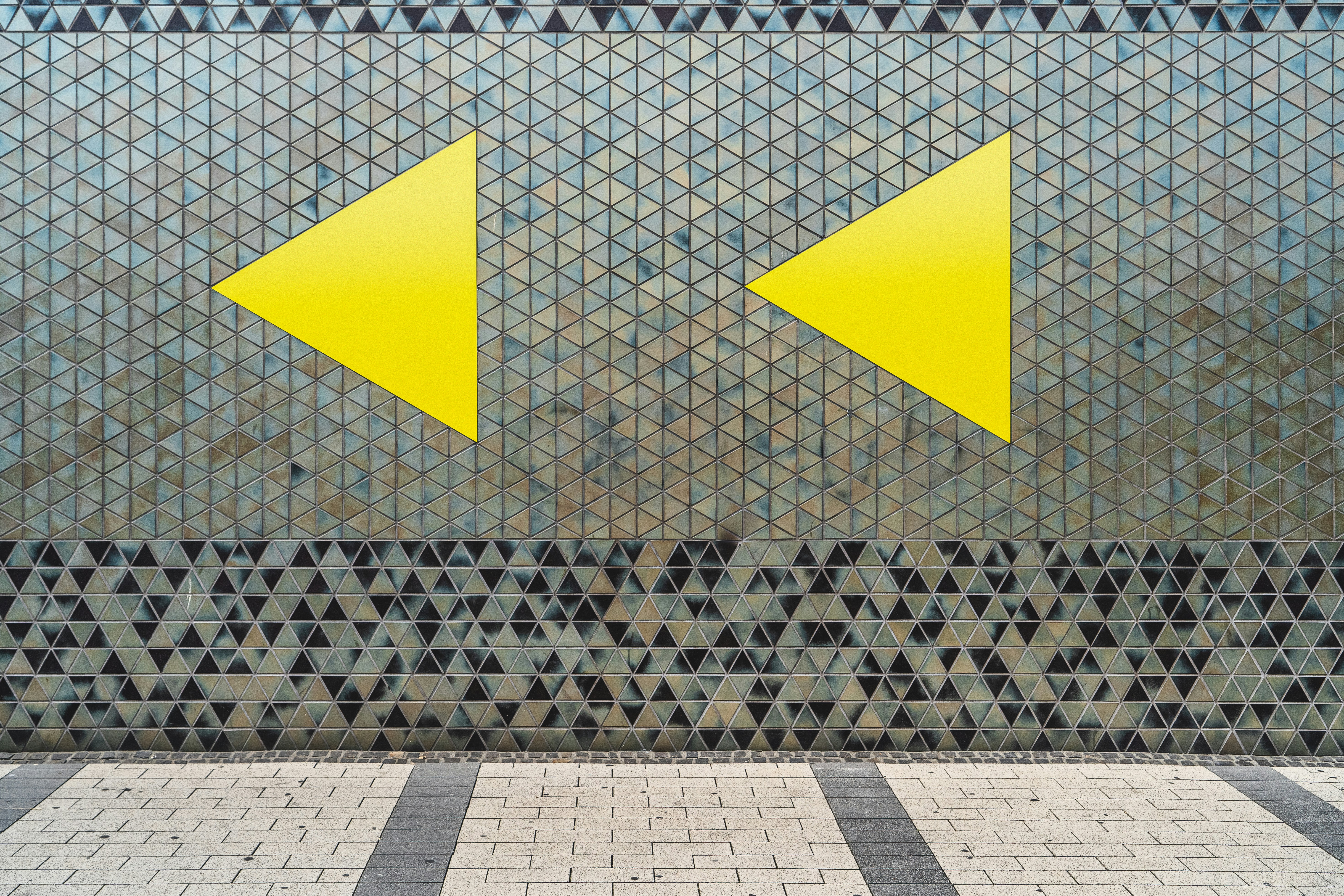 Two large yellow triangles juxtaposed against a textured, triangular-patterned wall create a striking visual contrast. The composition emphasizes geometric shapes and modern design.