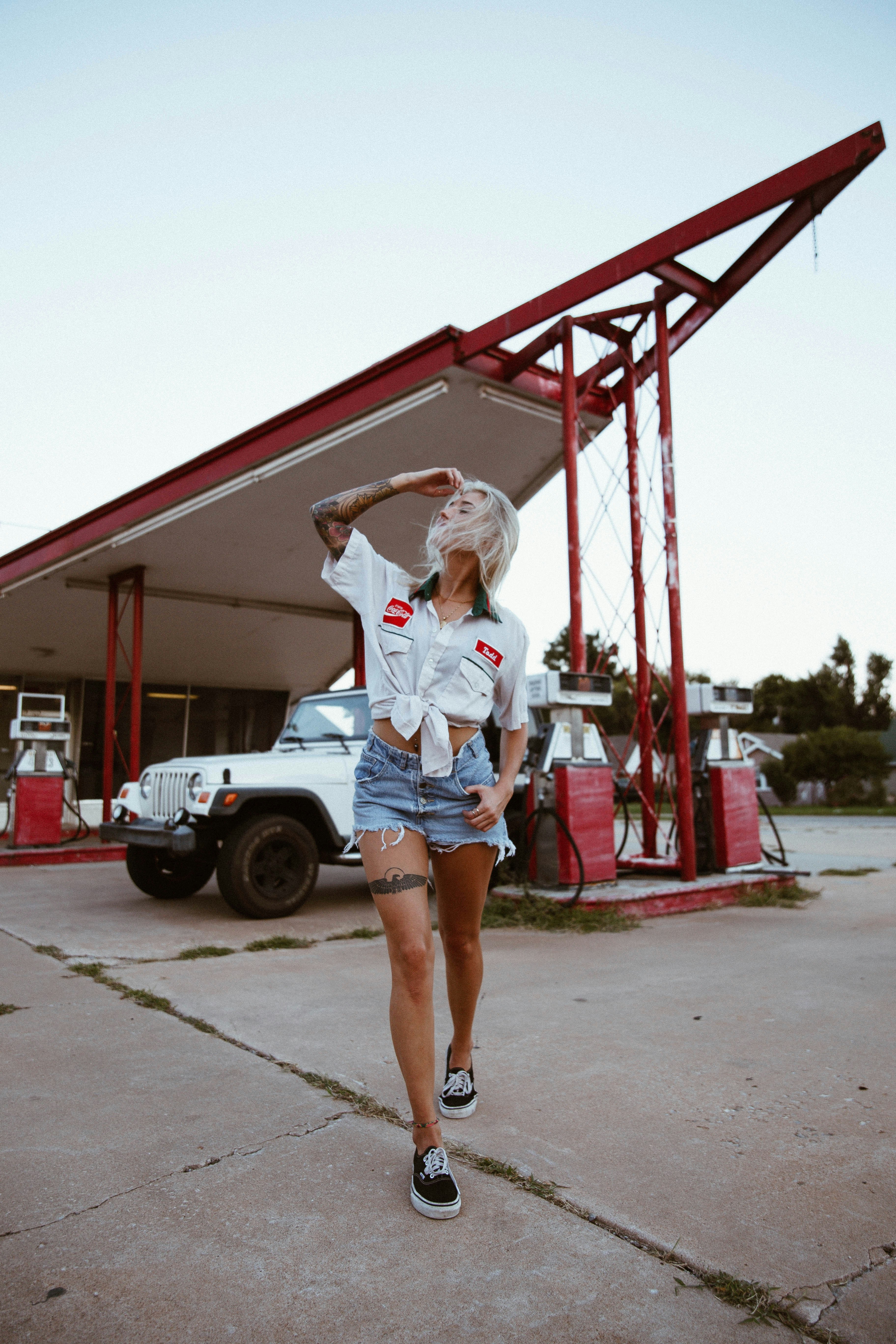 A young woman with a playful pose stands in front of a vintage gas station, showcasing her stylish outfit and tattoos. The classic jeep adds to the nostalgic atmosphere.