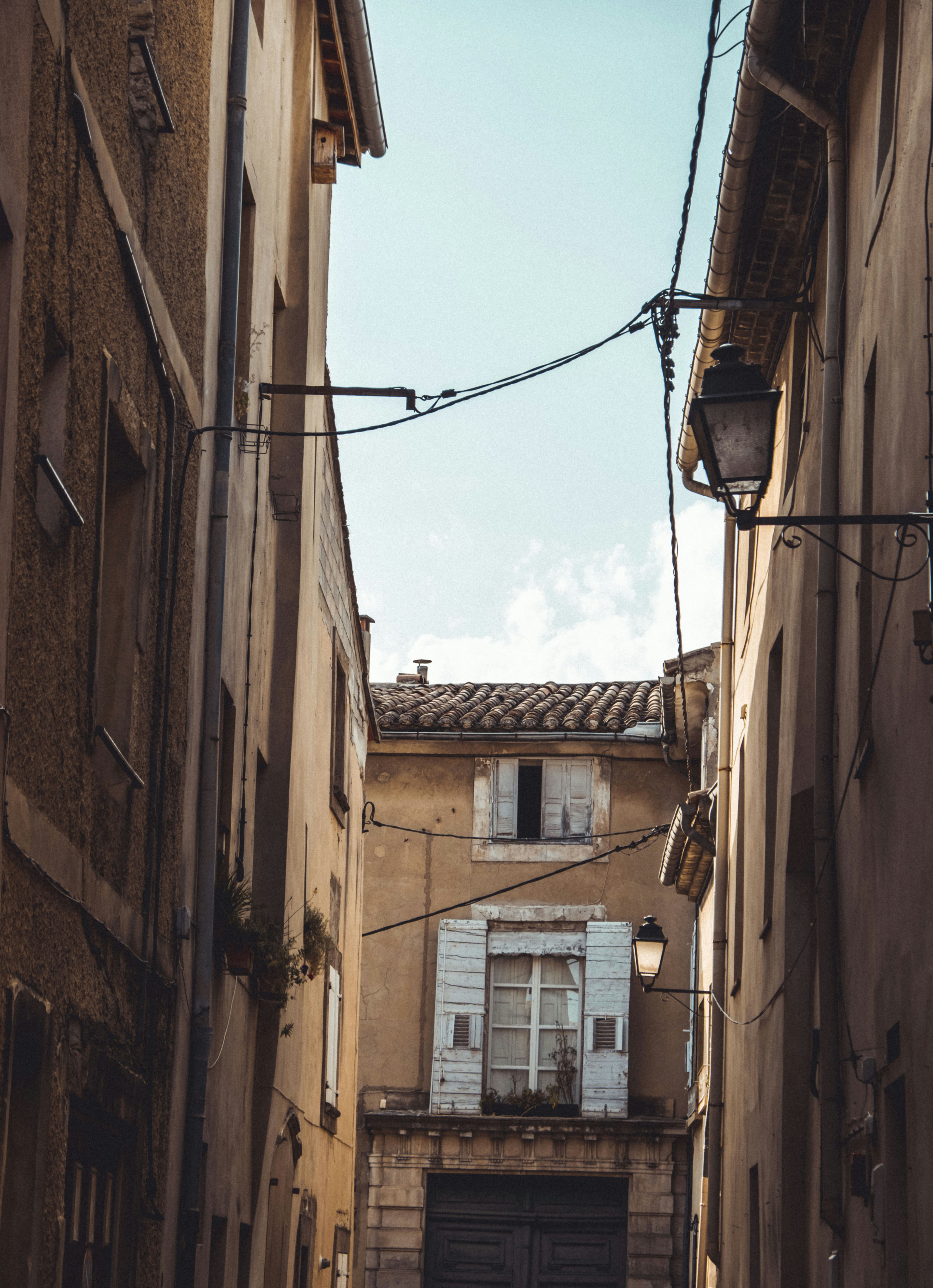 Charming alleyway framed by rustic buildings, showcasing a vintage facade with a window dressed in blue shutters. A street lamp adds to the quaint atmosphere.