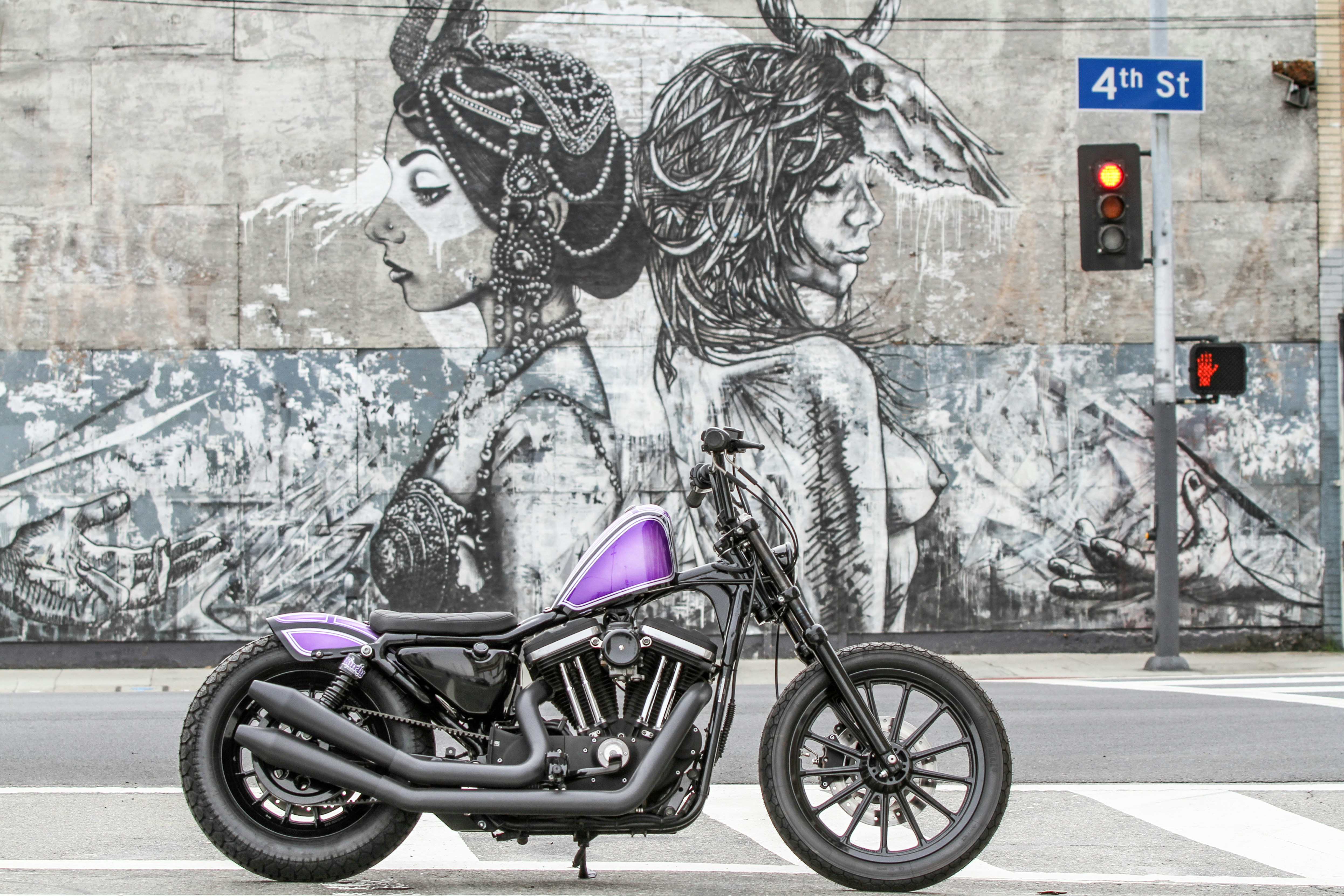 A custom motorcycle stands prominently against a vibrant street mural featuring two women, blending urban culture with artistic expression.
