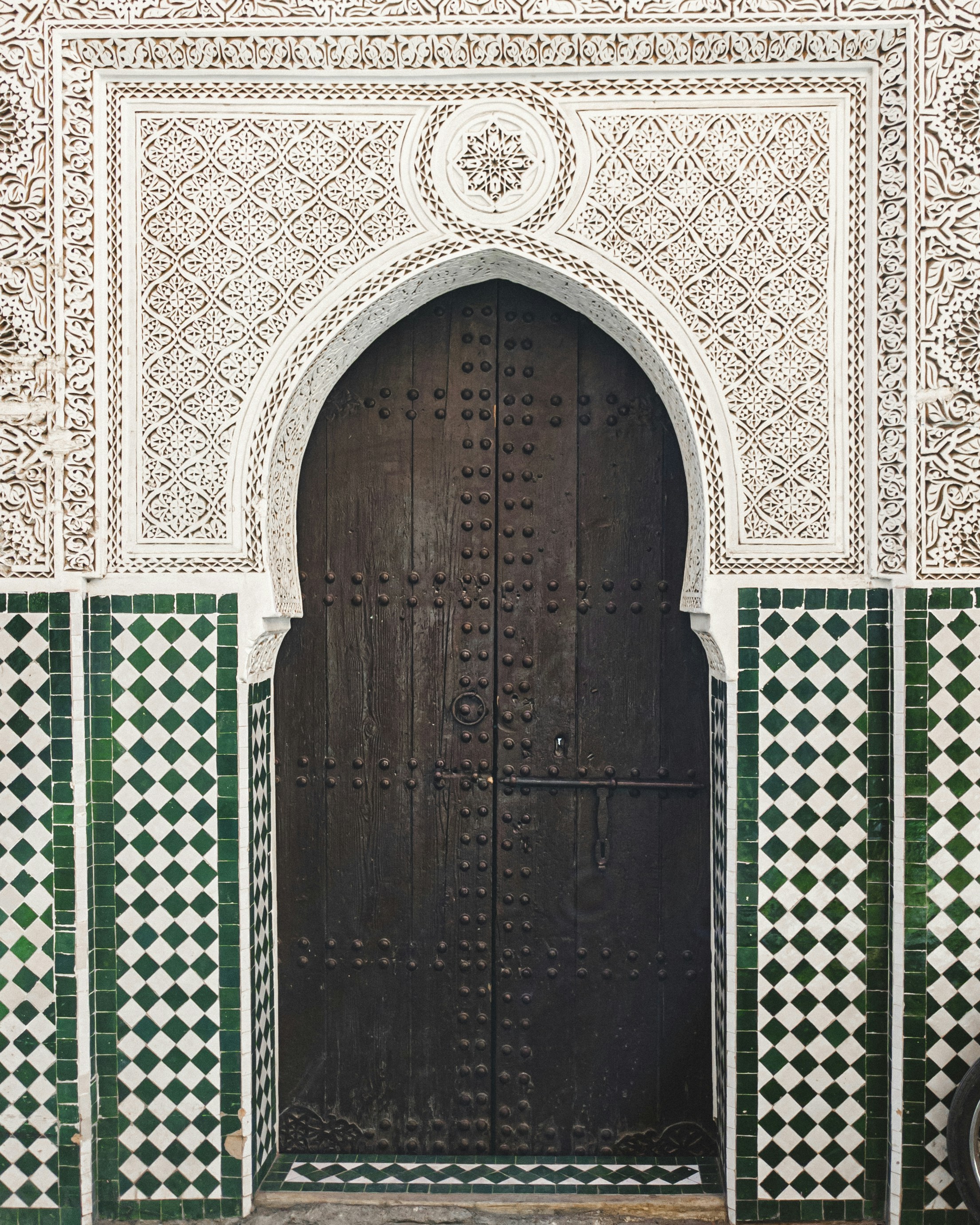 Elaborately carved archway framing a dark wooden door, flanked by vibrant green and white tiled patterns.