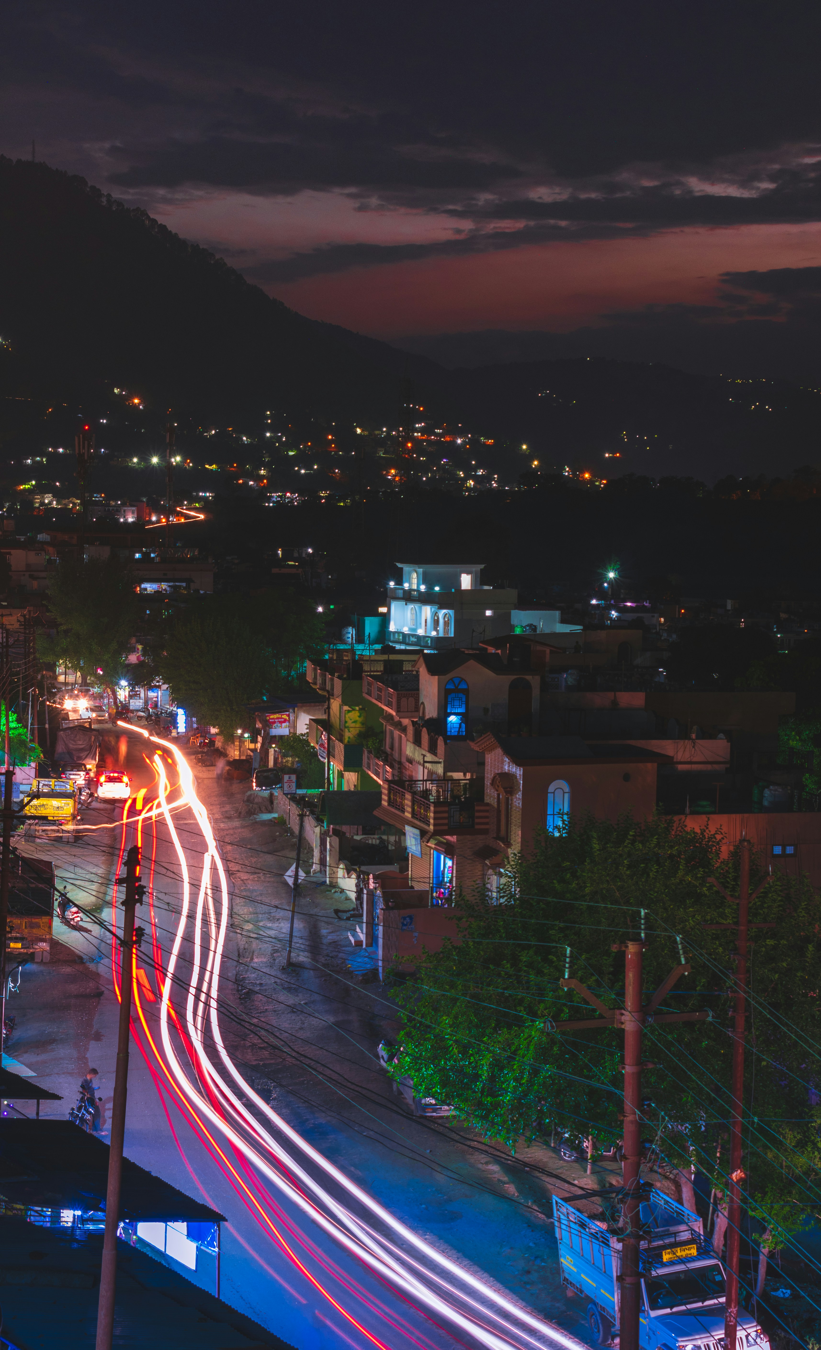 Vibrant street scene capturing light trails from vehicles on a bustling road at night, framed by illuminated buildings and a mountainous backdrop.