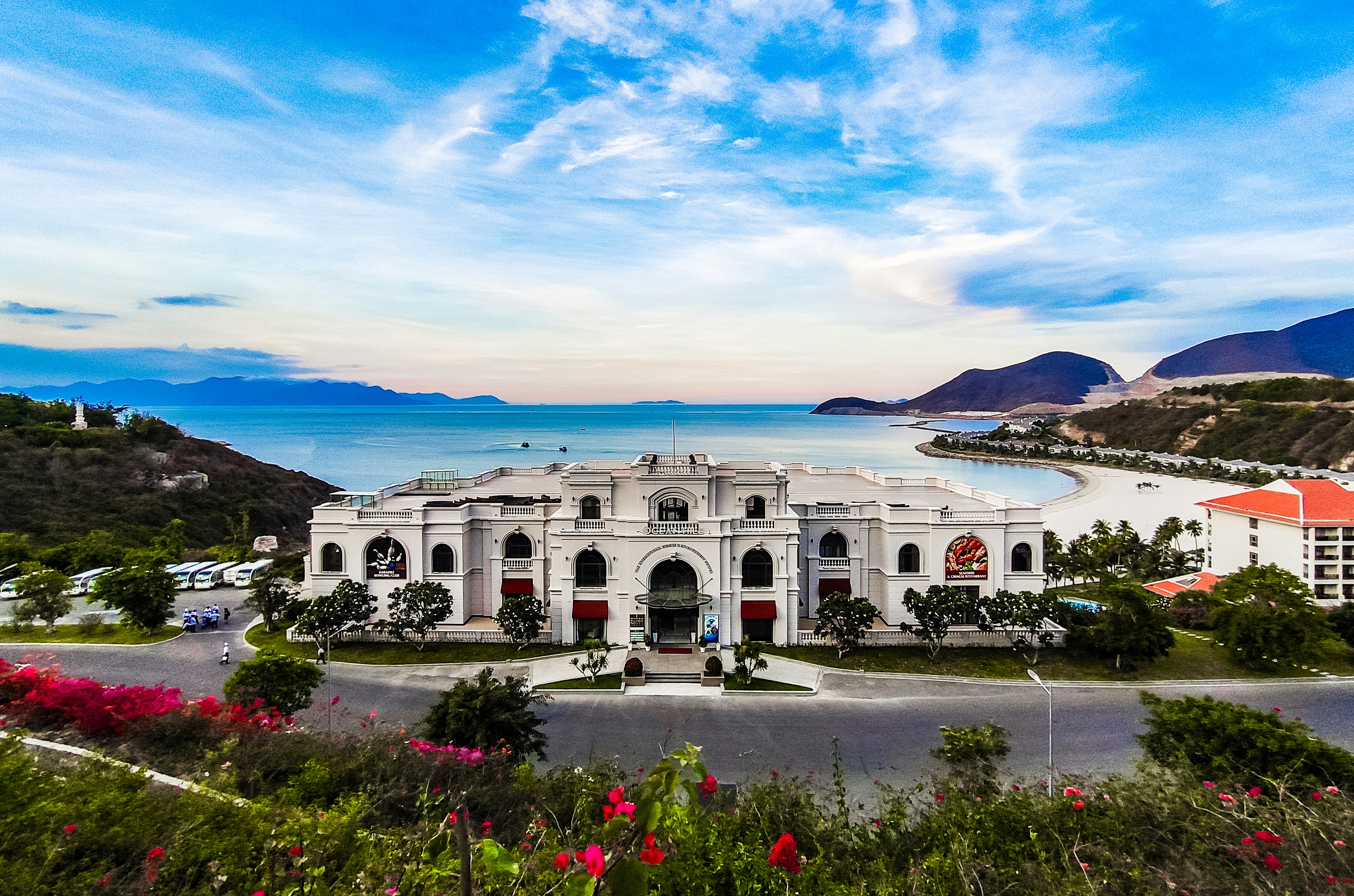 Grand white building overlooking a vibrant coastal landscape with mountains and sea under a vivid blue sky.