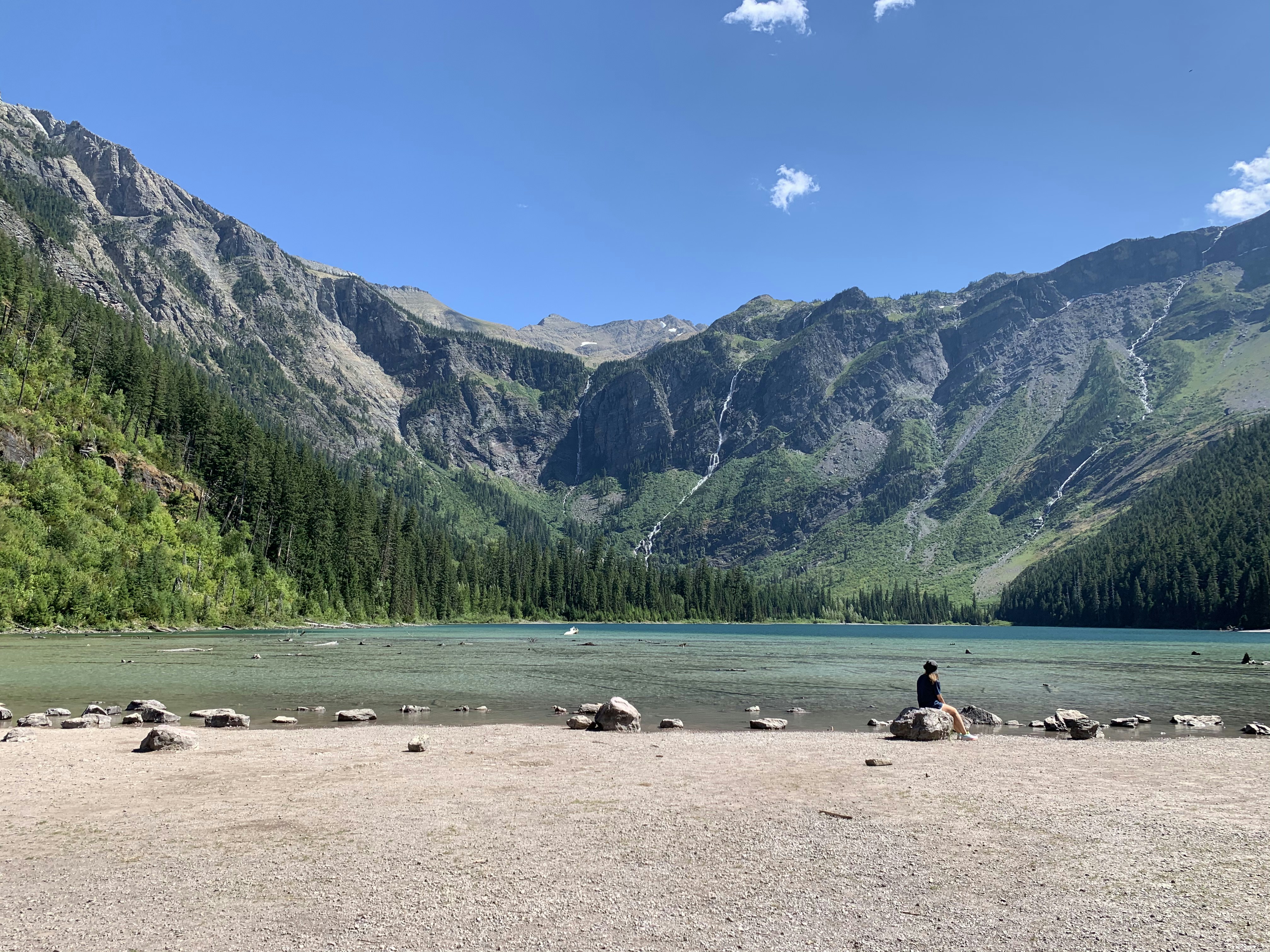 Person sitting by a serene lake surrounded by towering mountains and lush greenery under a clear blue sky.