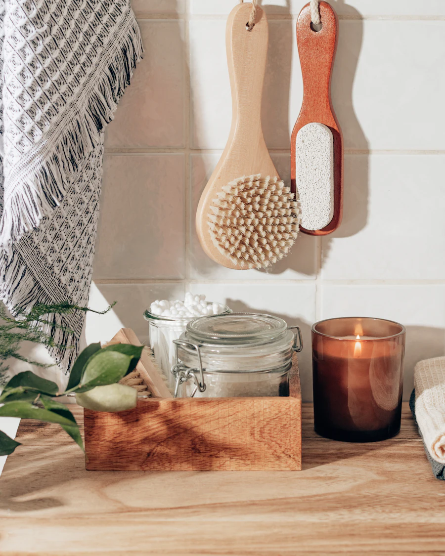 Bathroom shelf still life with hanging wooden body brushes, a lit amber candle, glass jars, and a woven cloth against white tile. Soft natural light creates warm shadows and a cozy, clean feel.