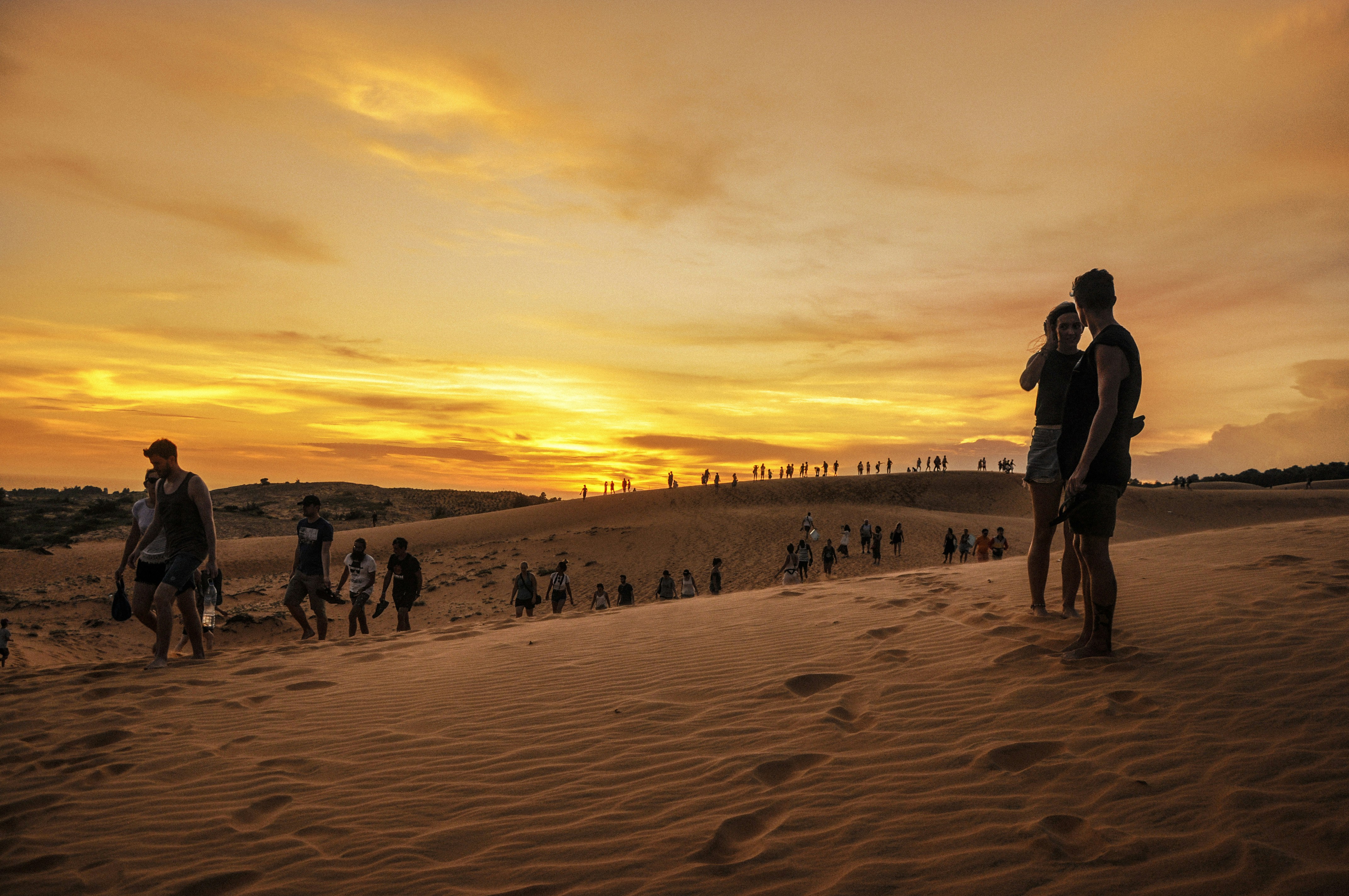 Silhouetted figures traverse the undulating dunes as the sun sets, casting a warm glow across the sandy landscape.