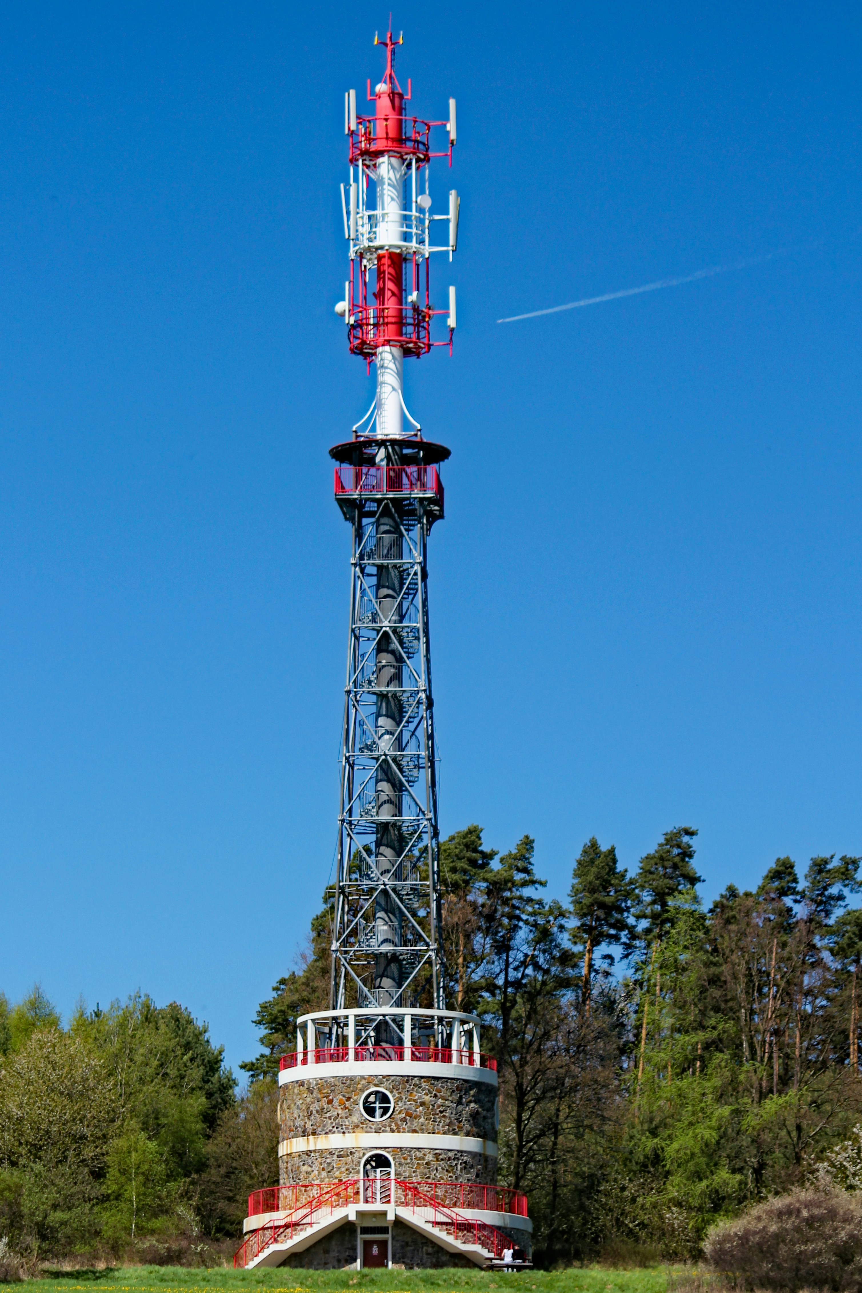 Telecommunication tower with a striking red and white design, surrounded by lush greenery under a clear blue sky.