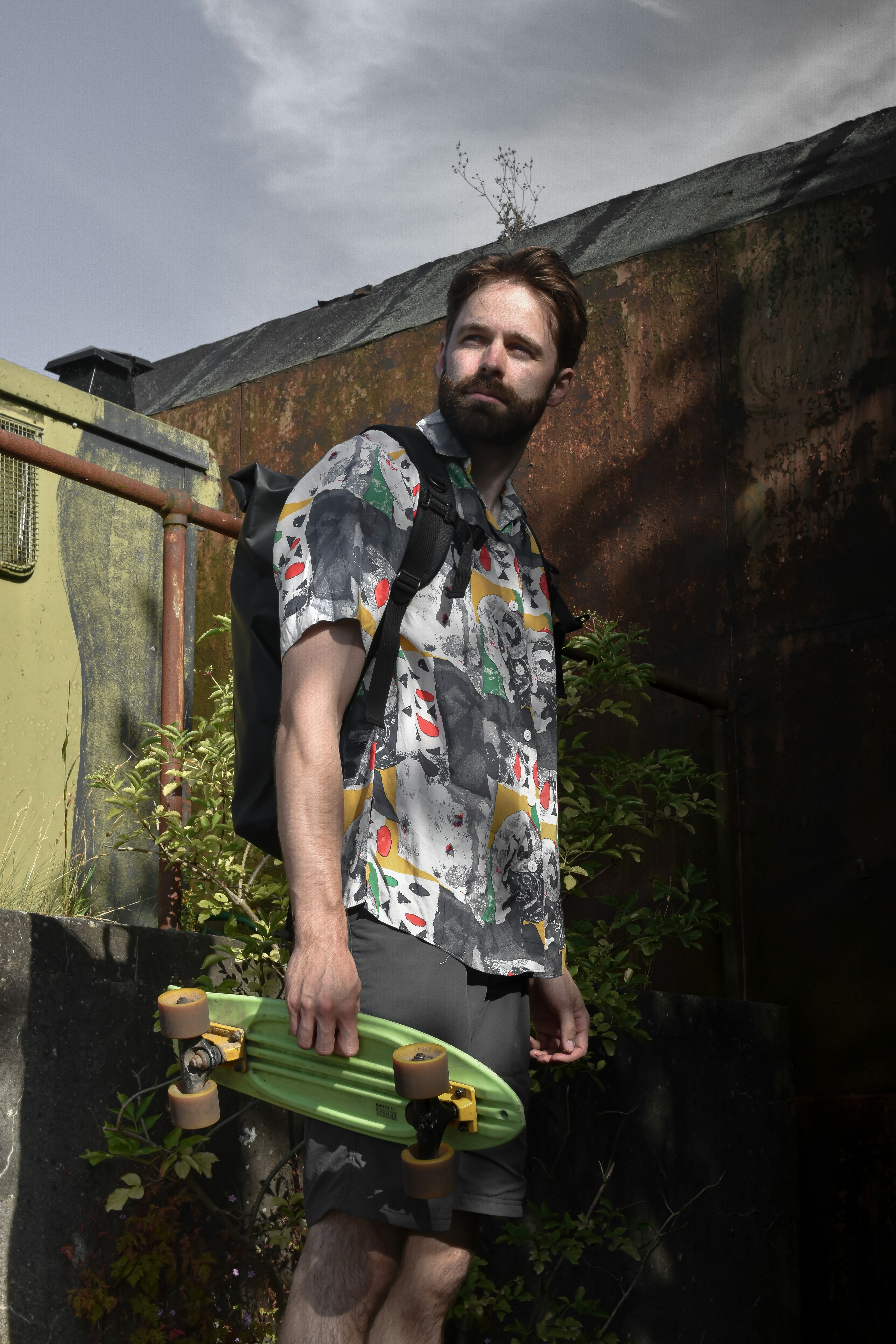 A young man stands confidently with a skateboard in hand, surrounded by urban decay and vibrant greenery. His colorful shirt contrasts with the weathered backdrop.
