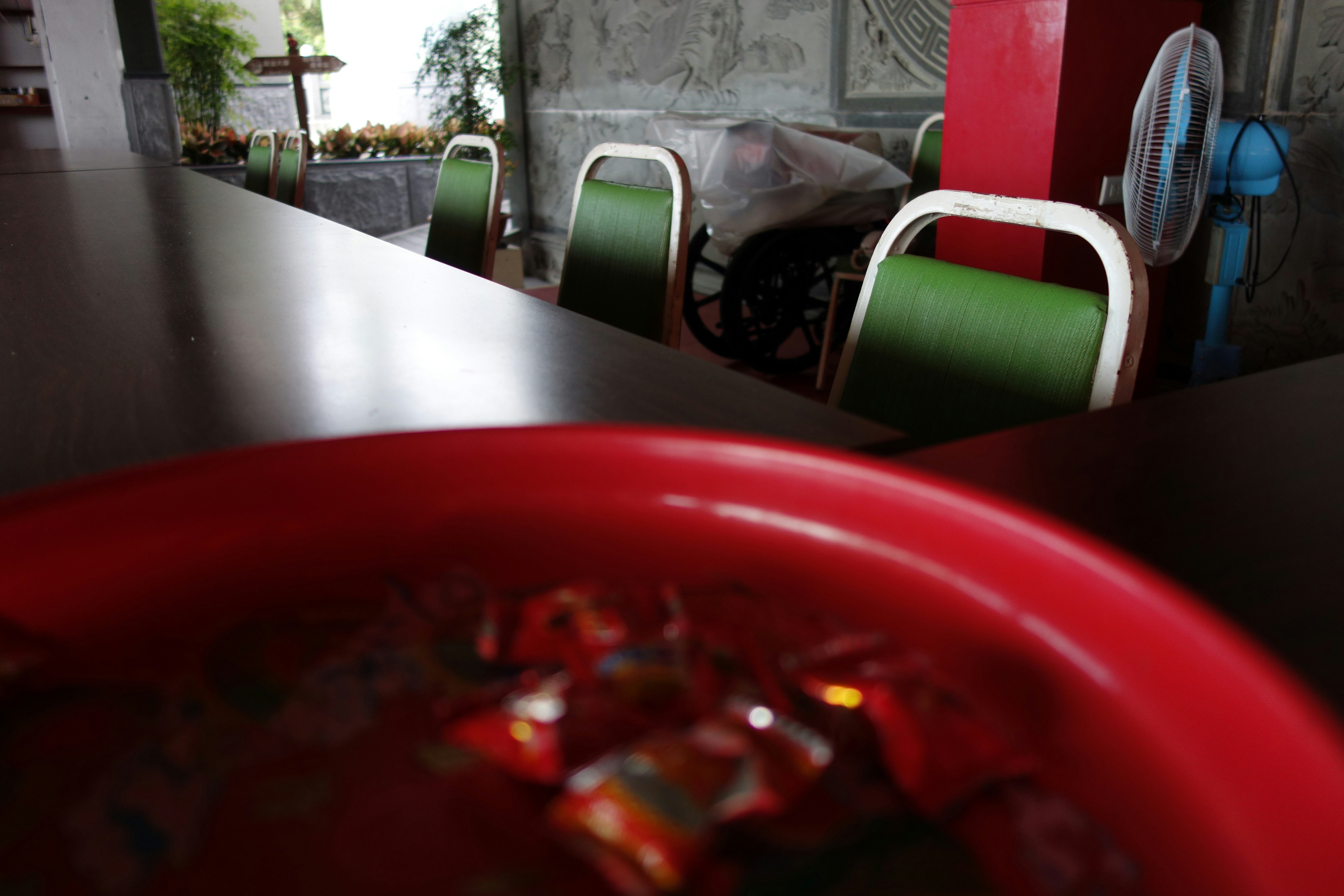 Crimson Bowl at the Edge: A Dim Café Scene with Green Chairs and a Blue Fan