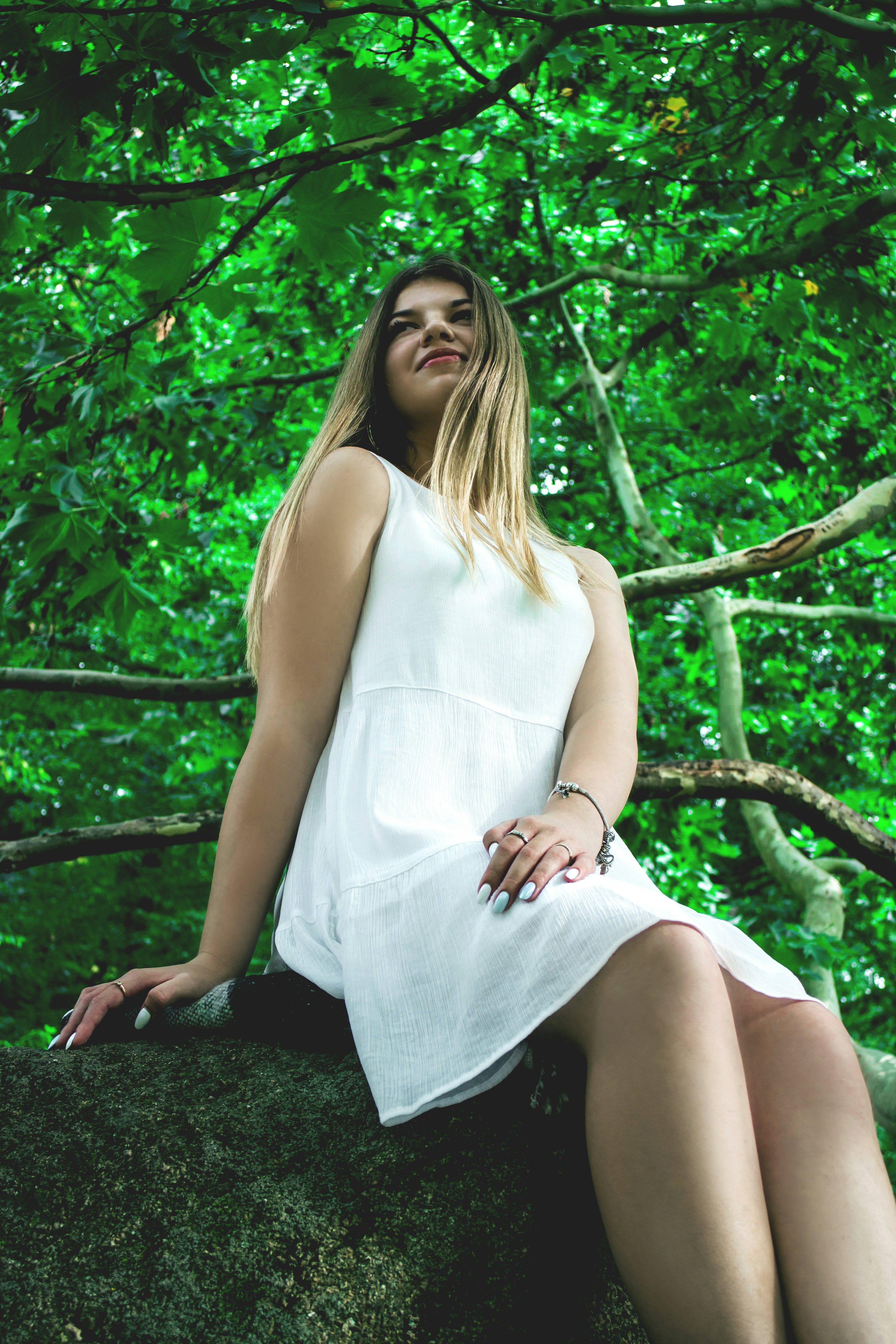 A young woman in a white dress sits gracefully on a moss-covered rock, surrounded by lush green foliage, embodying tranquility and connection to nature.