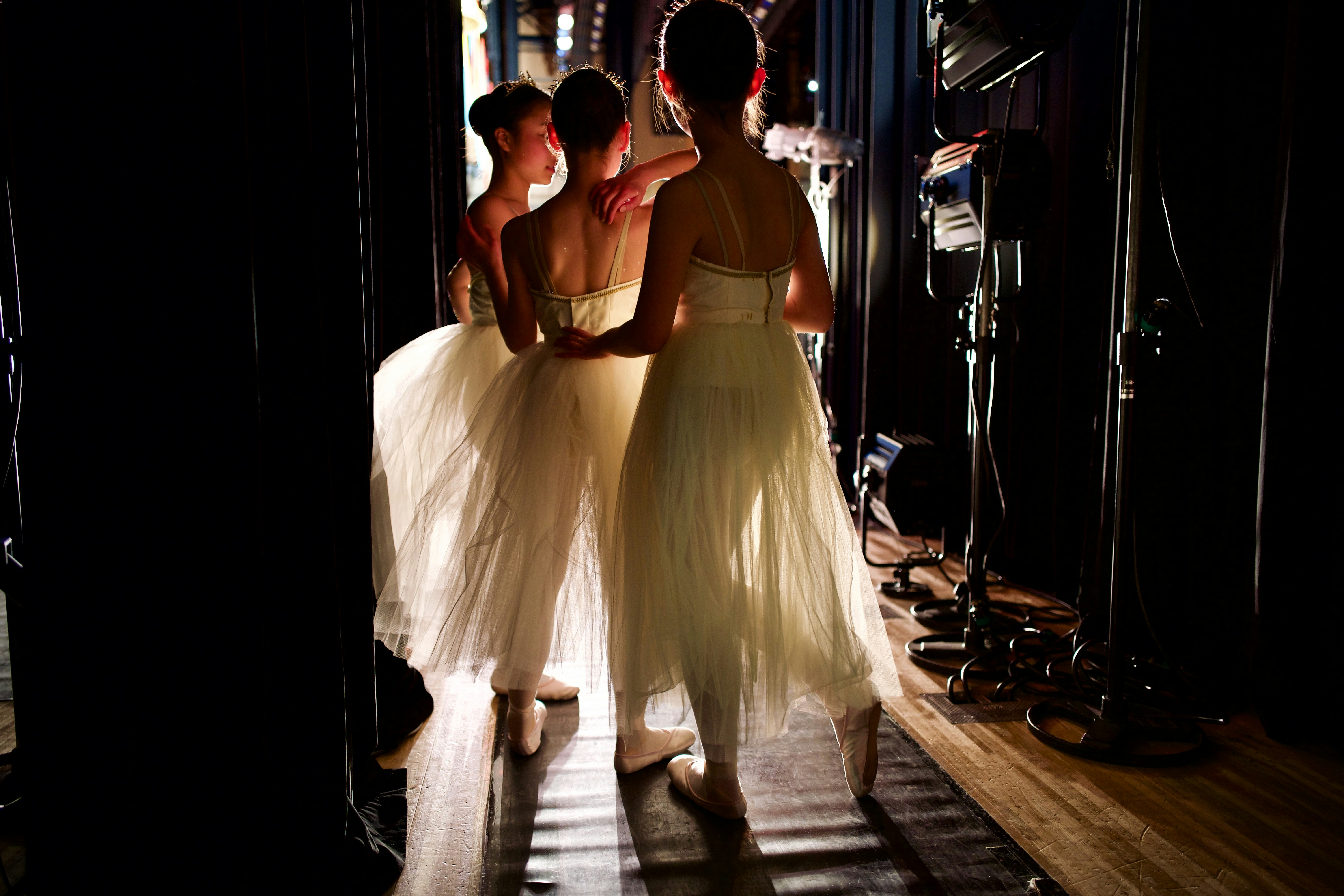 Three ballet dancers in elegant tutus share a moment backstage, illuminated by soft lighting as they prepare to take the stage.