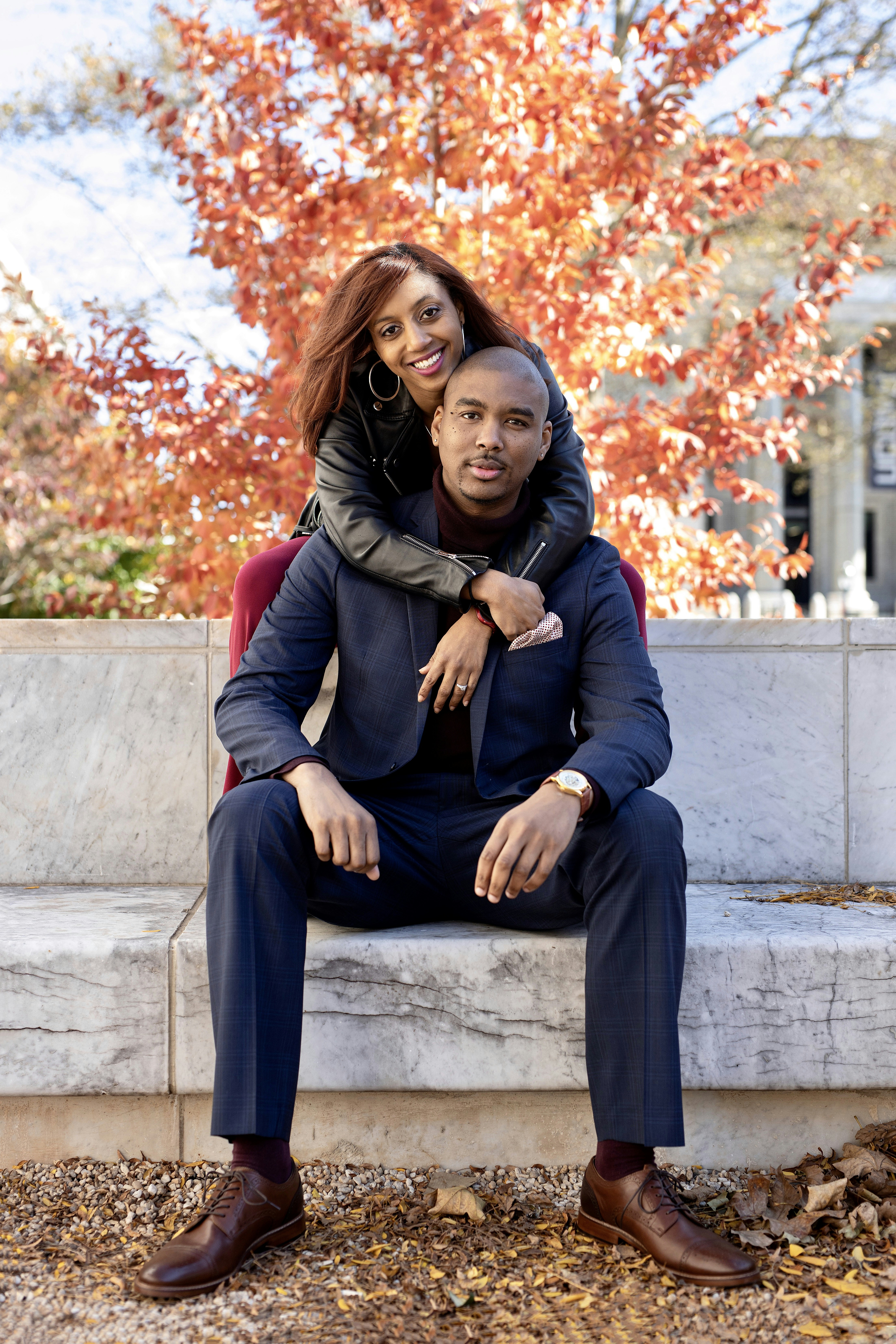 Couple seated on a marble bench surrounded by vibrant autumn foliage, exuding warmth and connection. The backdrop features bright orange leaves, enhancing the intimate atmosphere.