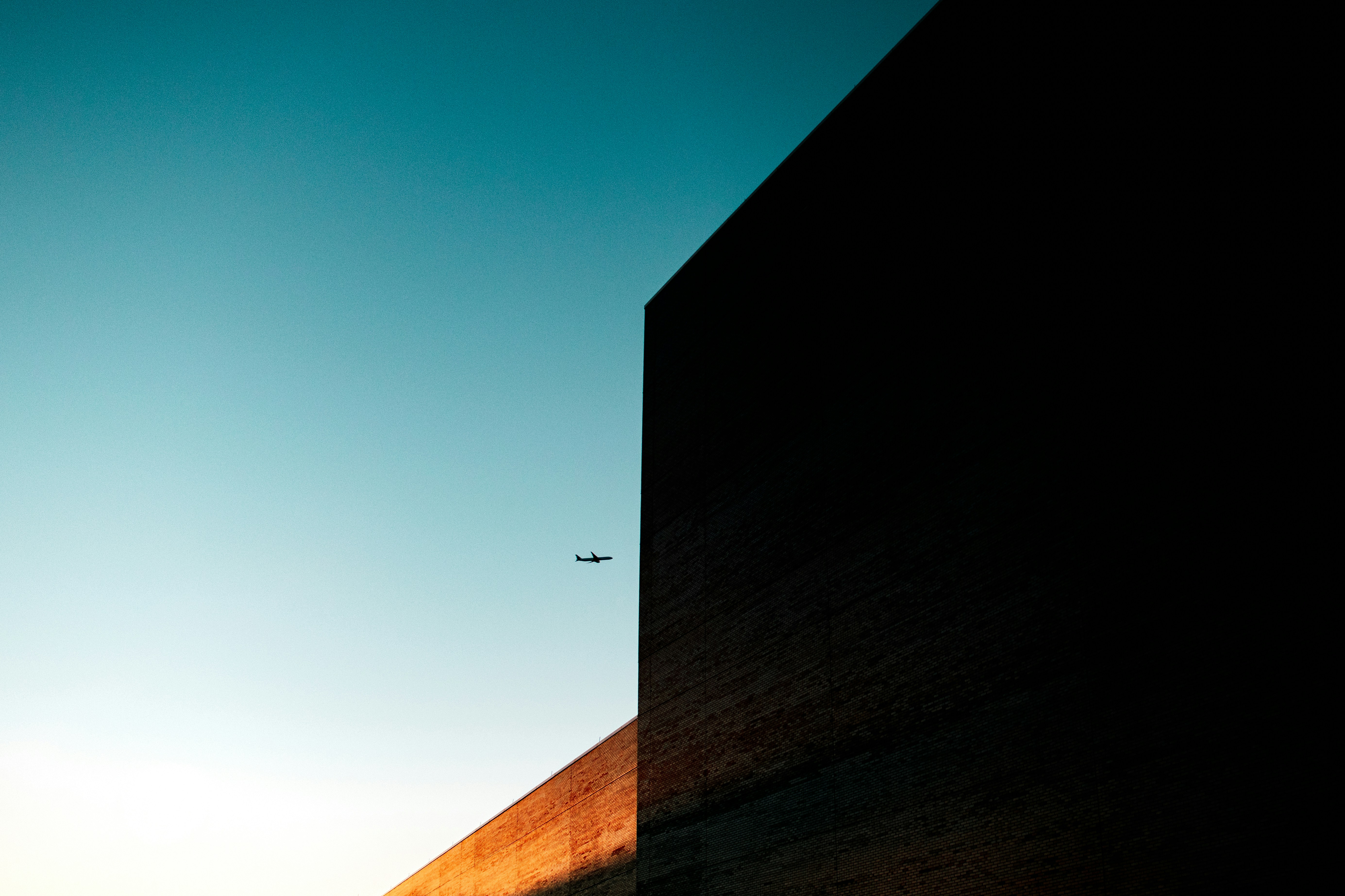 A sleek airplane glides through a clear sky, framed by the stark geometric lines of a modern architectural structure. The warm glow of sunset contrasts with the cool tones above.
