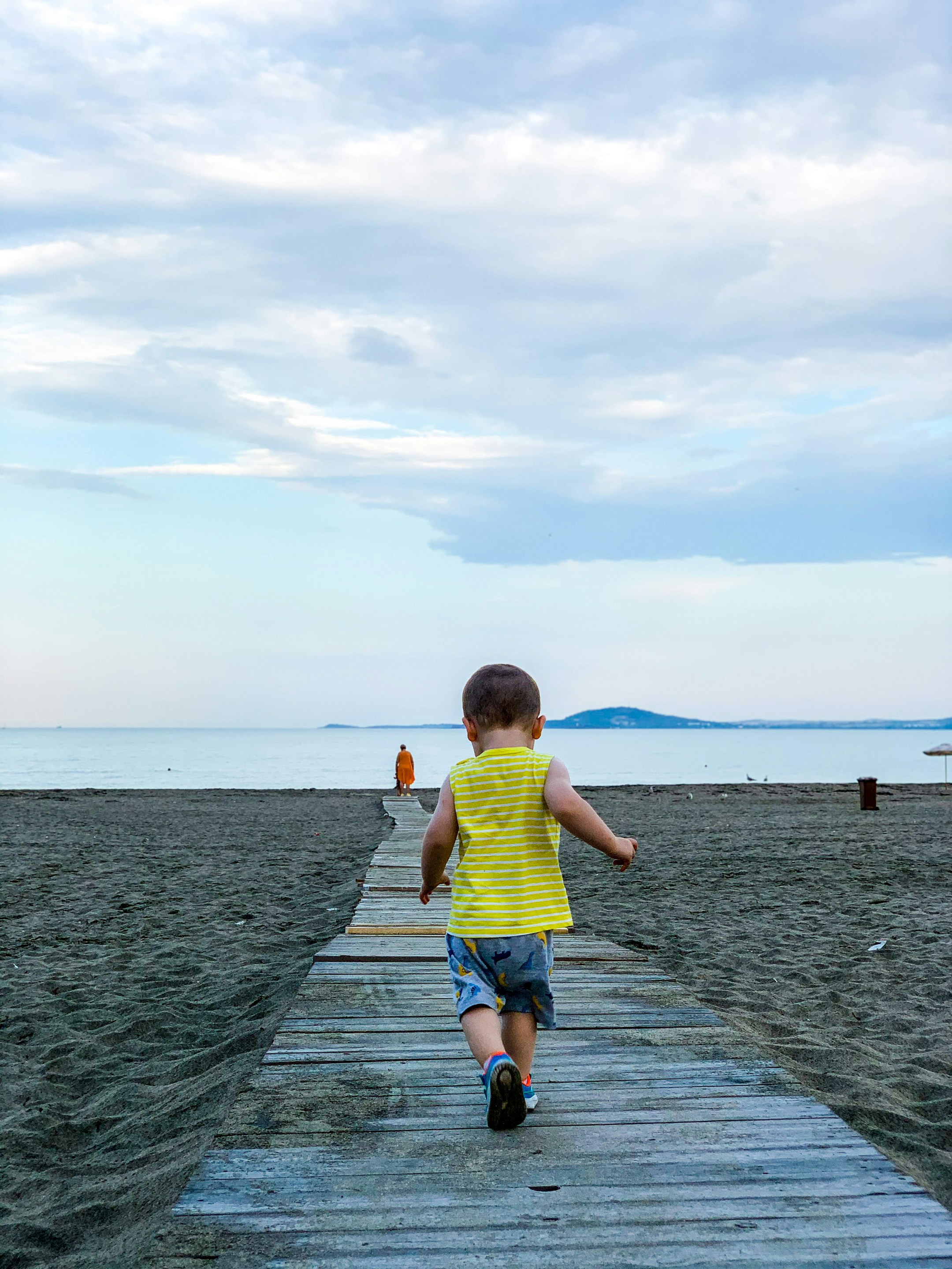 A young boy in a yellow tank top runs along a wooden pathway leading to the sea, with a figure in the distance silhouetted against the water. The scene captures a moment of exploration and freedom.