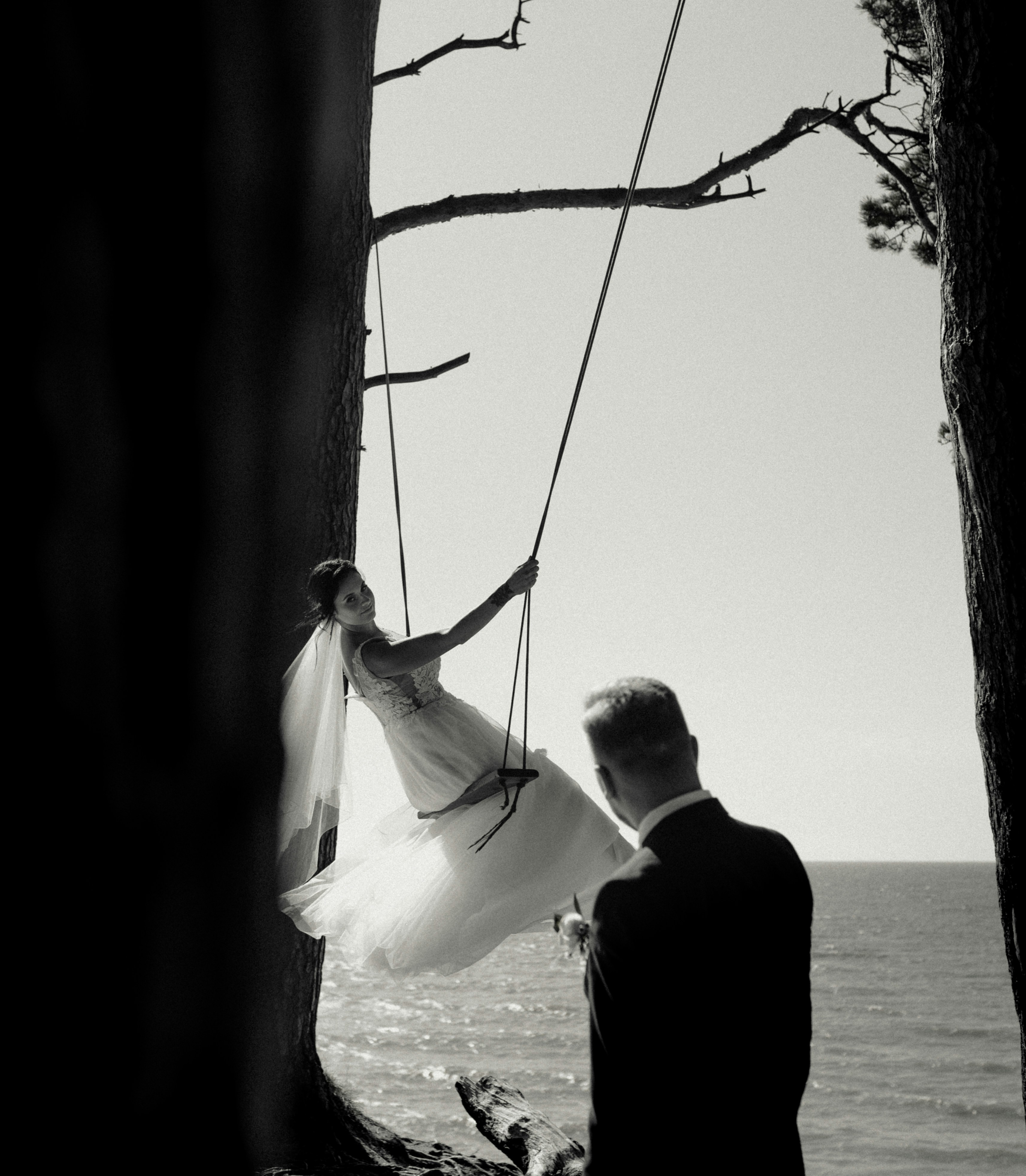 Bride on a swing suspended from a tree, gazing at her groom by the shore. The scene captures a playful moment amidst a serene backdrop.