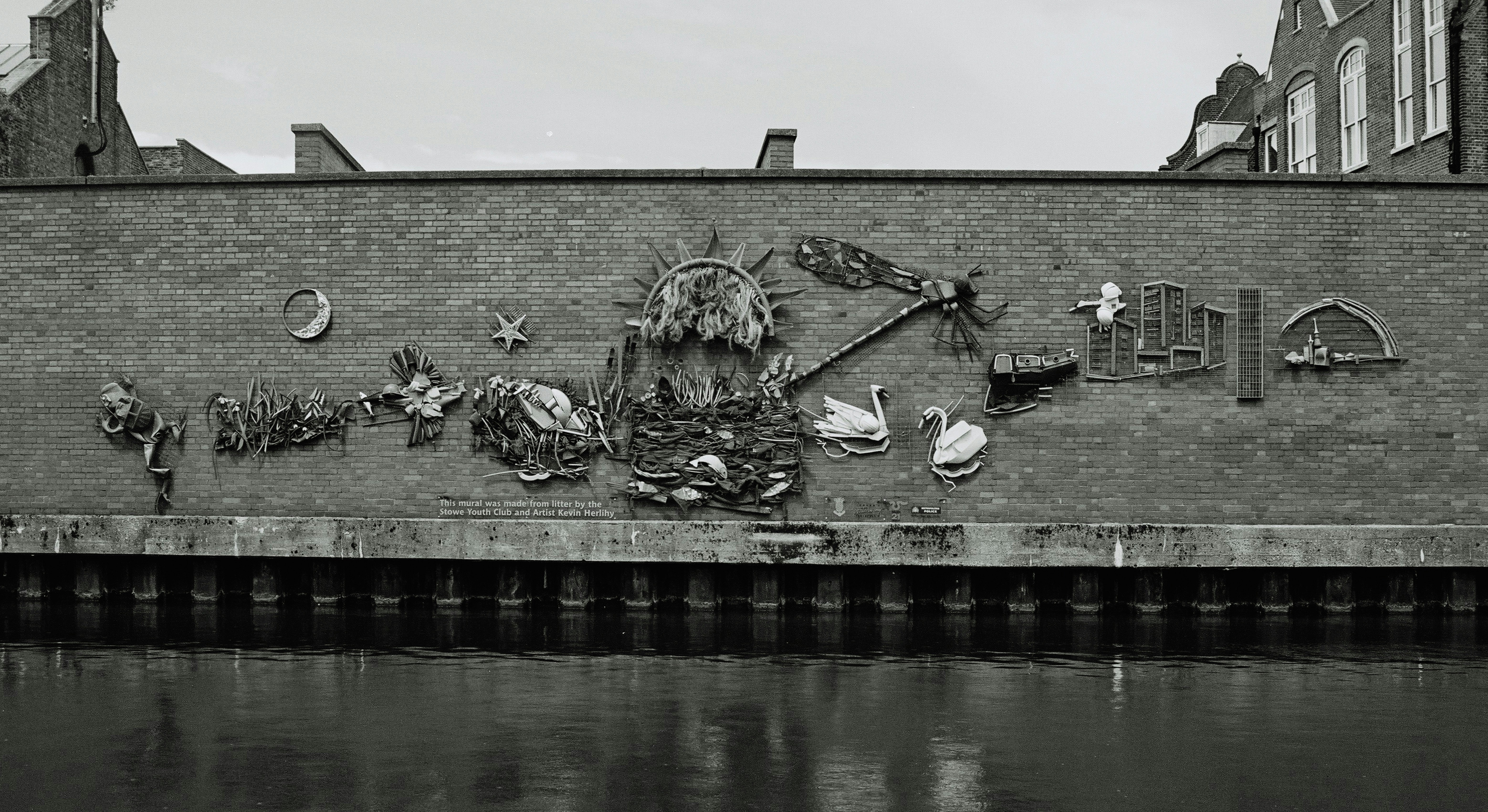 Monochrome mural of natural elements on a brick wall beside a canal, blending art with urban landscape.