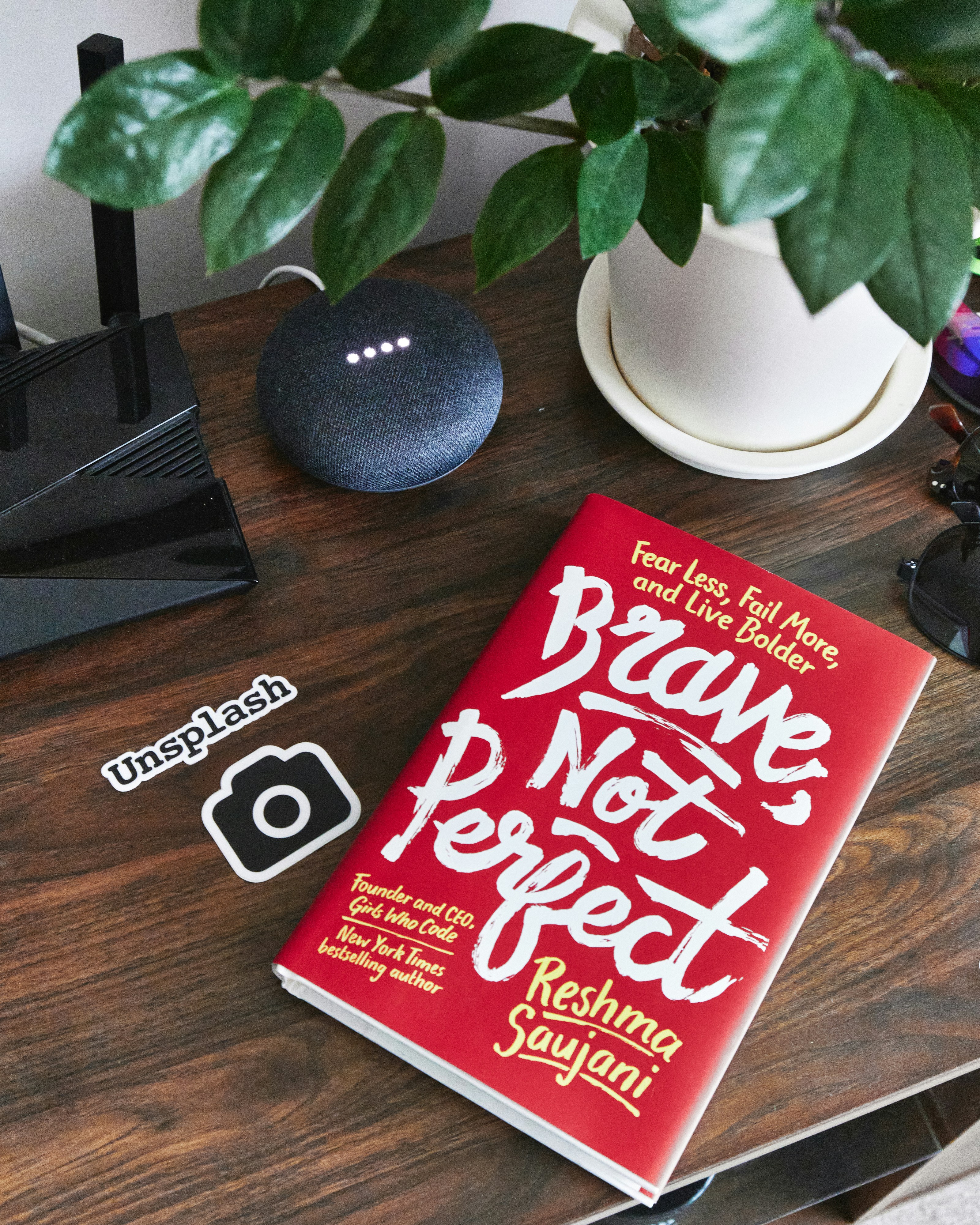 A vibrant book titled 'Brave, Not Perfect' by Reshma Saujani rests on a wooden desk alongside a smart speaker and a potted plant.