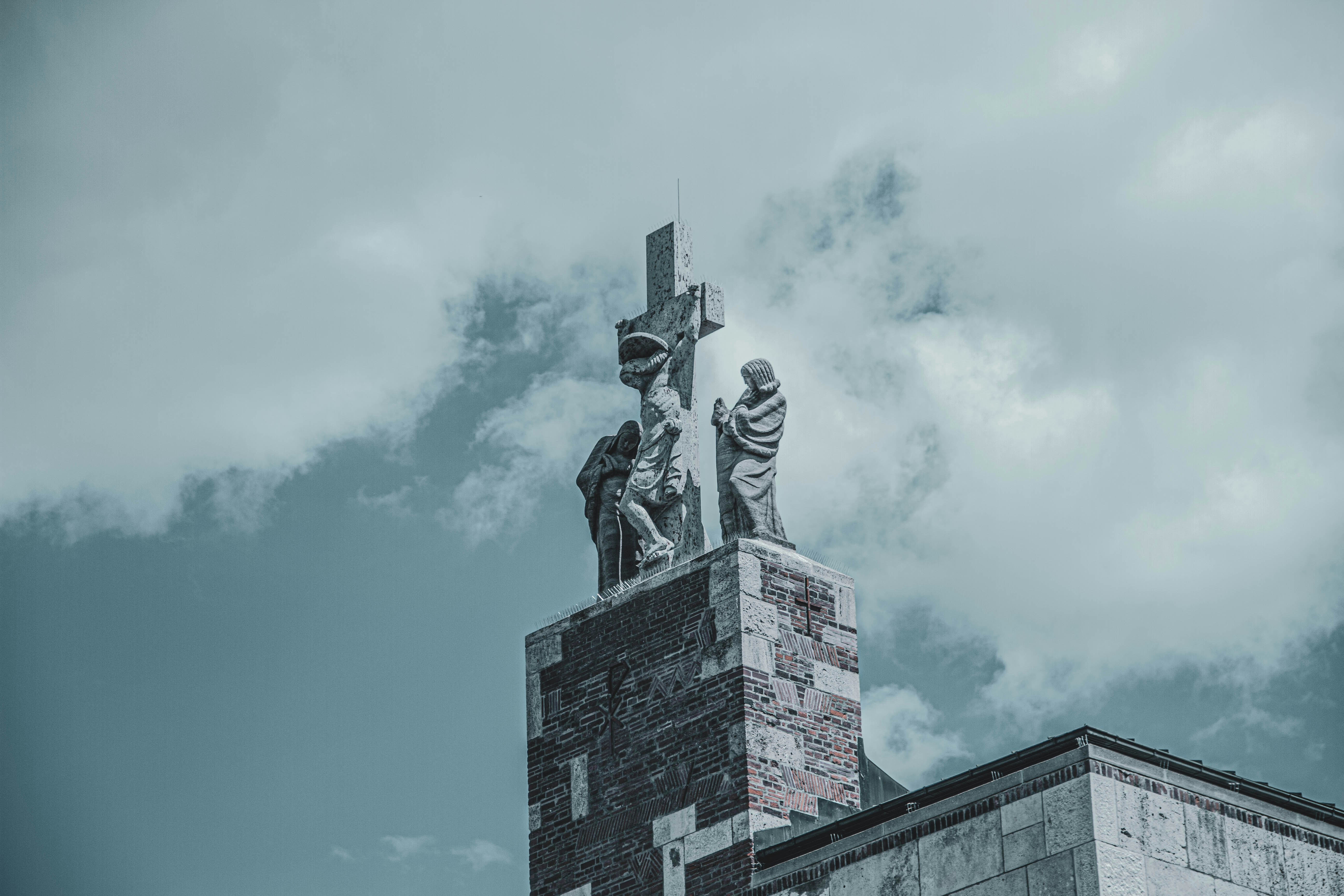 Statues of two figures beside a cross atop a building, set against a cloudy sky. The scene evokes a sense of reverence and history.