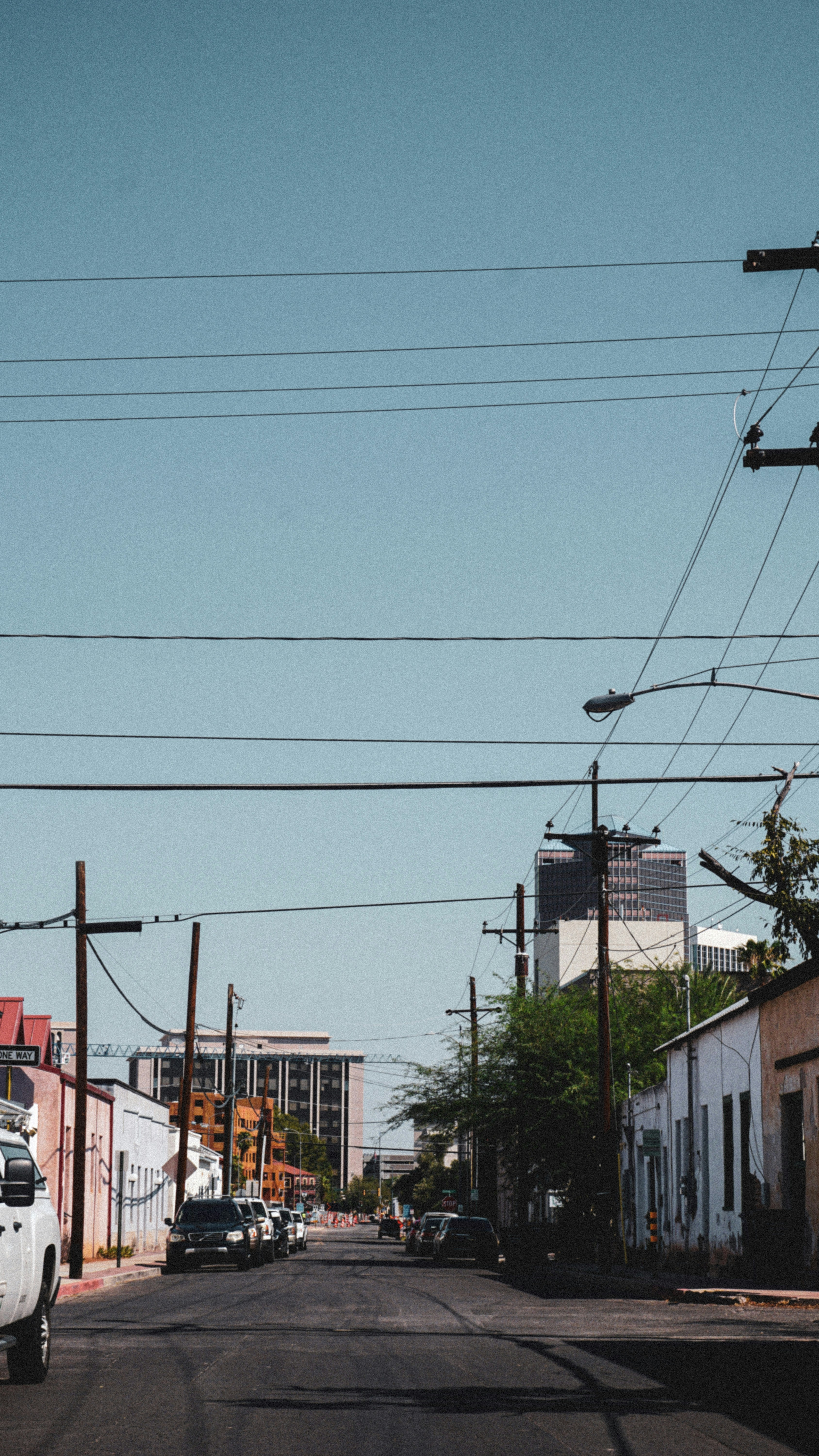 A sunlit urban street scene featuring parked vehicles and power lines, framed by city buildings in the background.