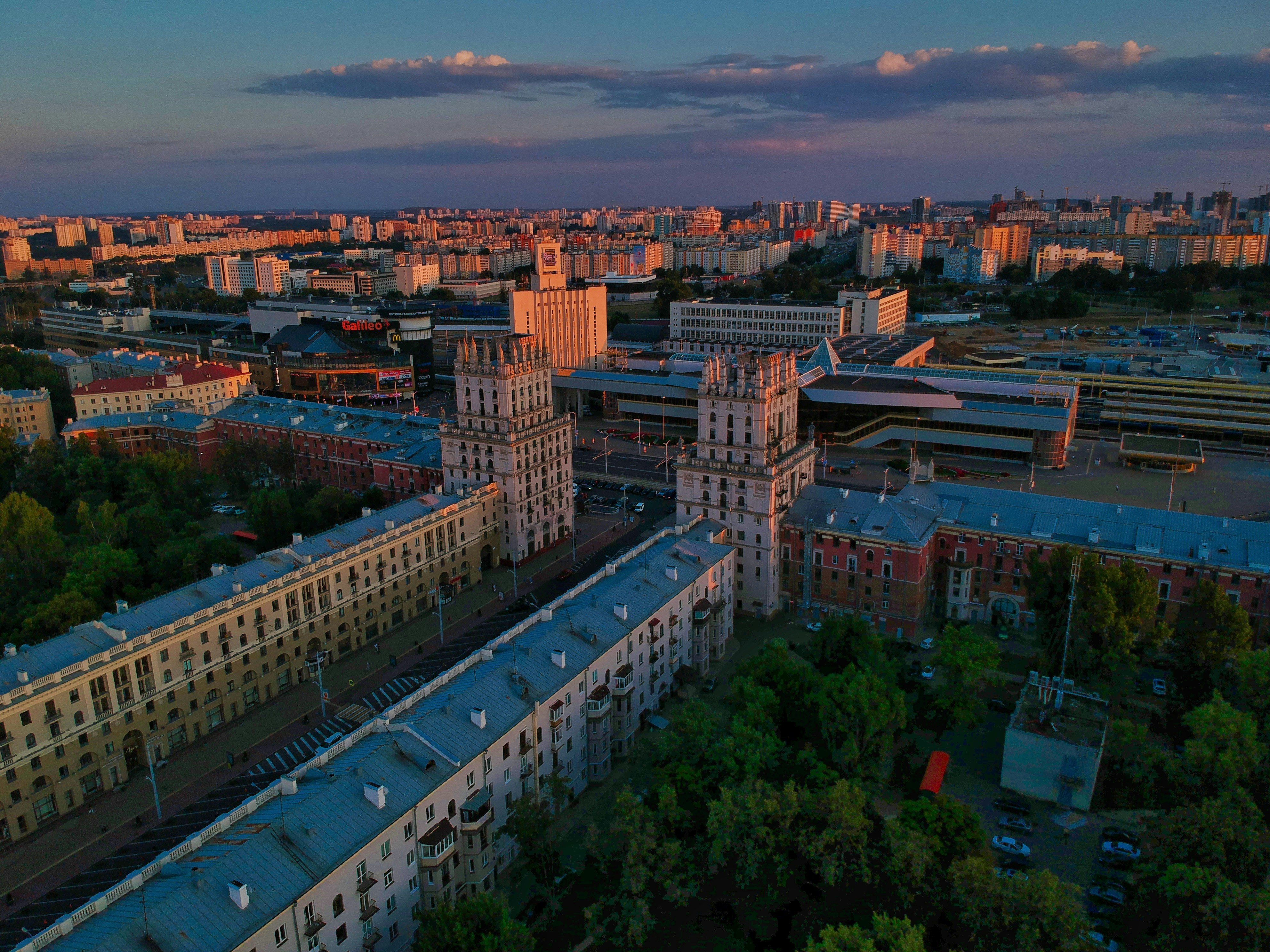 Cityscape at sunset featuring a central, symmetrical building surrounded by urban sprawl and greenery under a colorful sky.