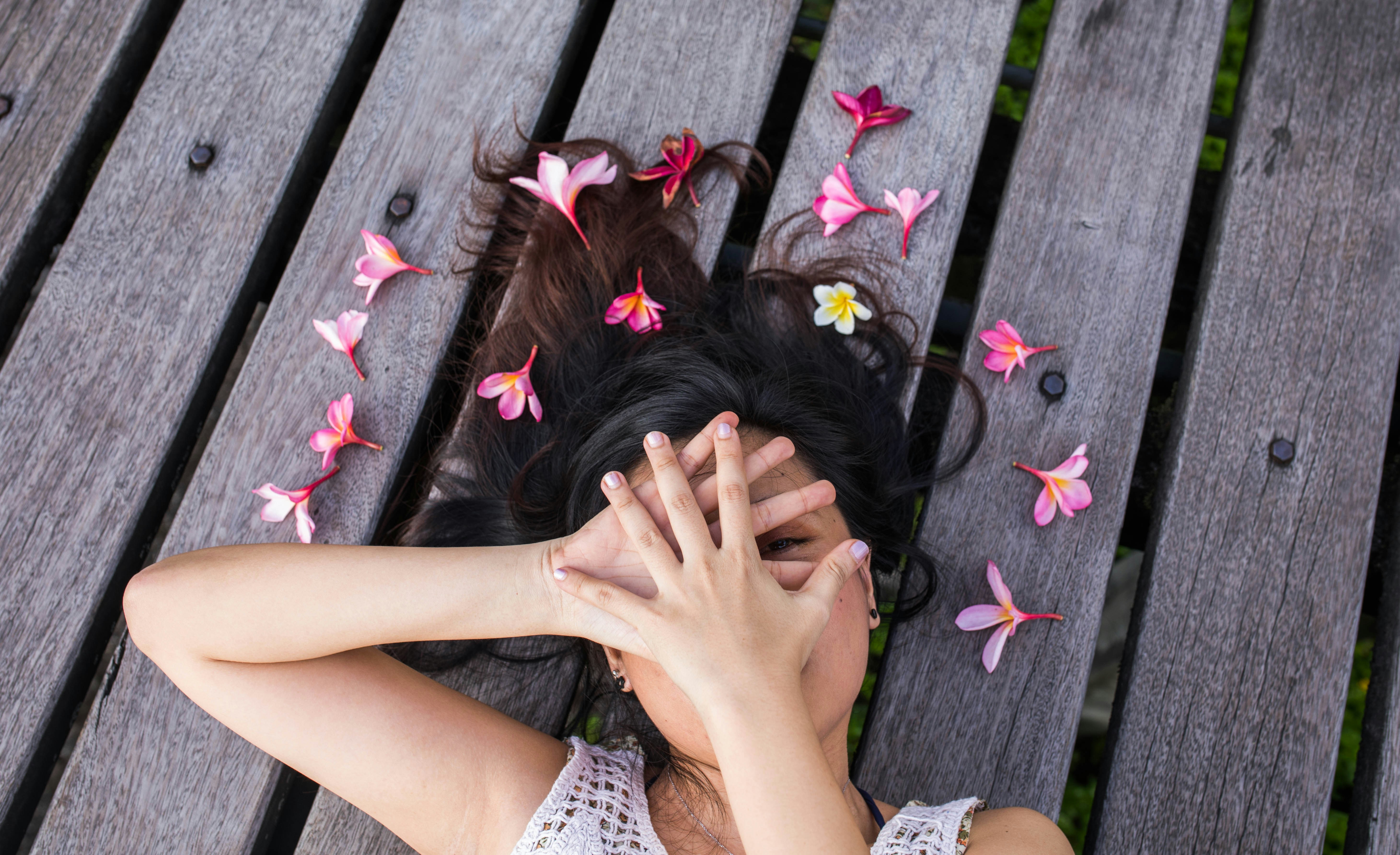Woman lying on wooden boards with hands covering face, surrounded by pink and white petals.