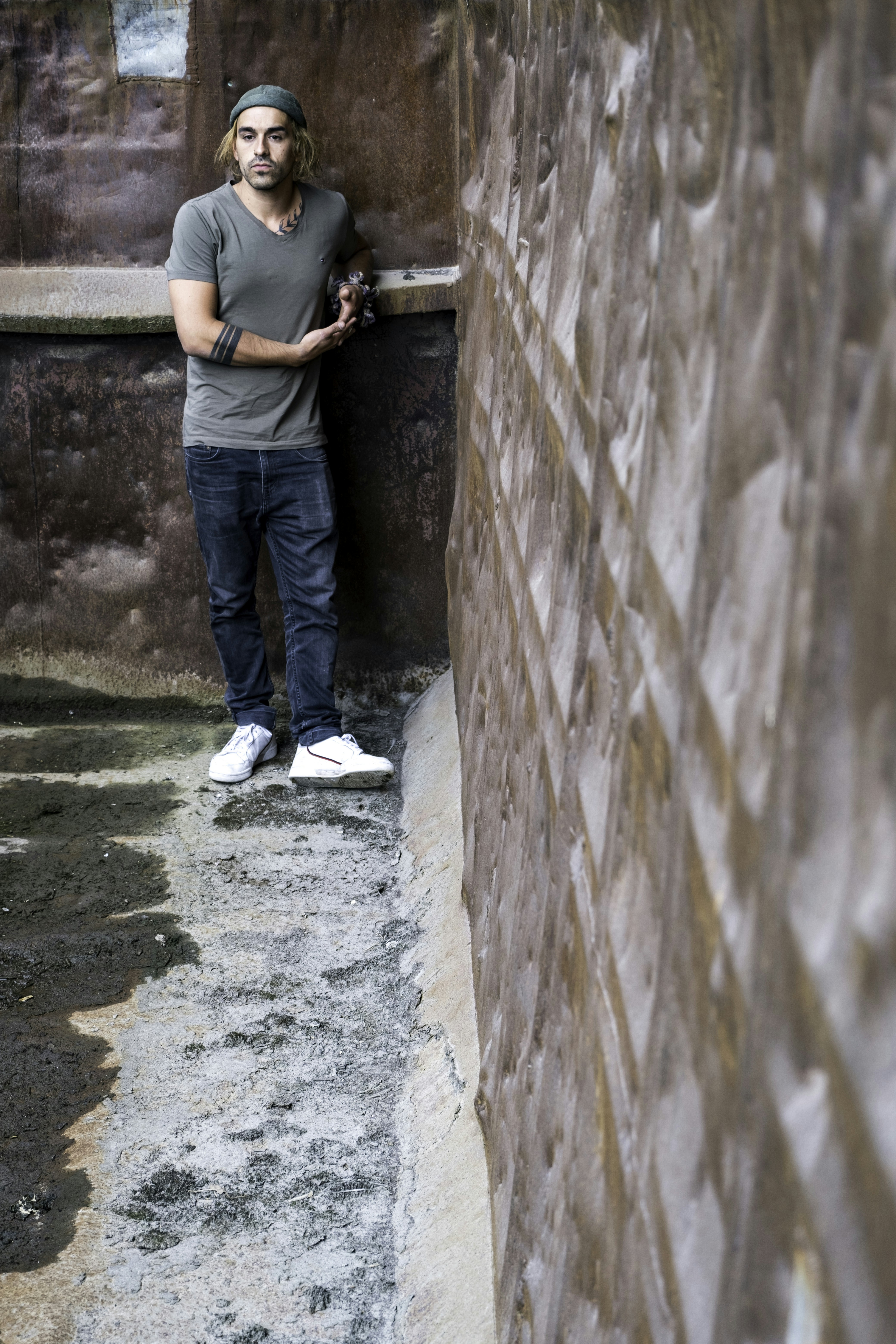 A man stands against a textured wall, holding a small object, immersed in thought. The setting exudes a sense of urban isolation.