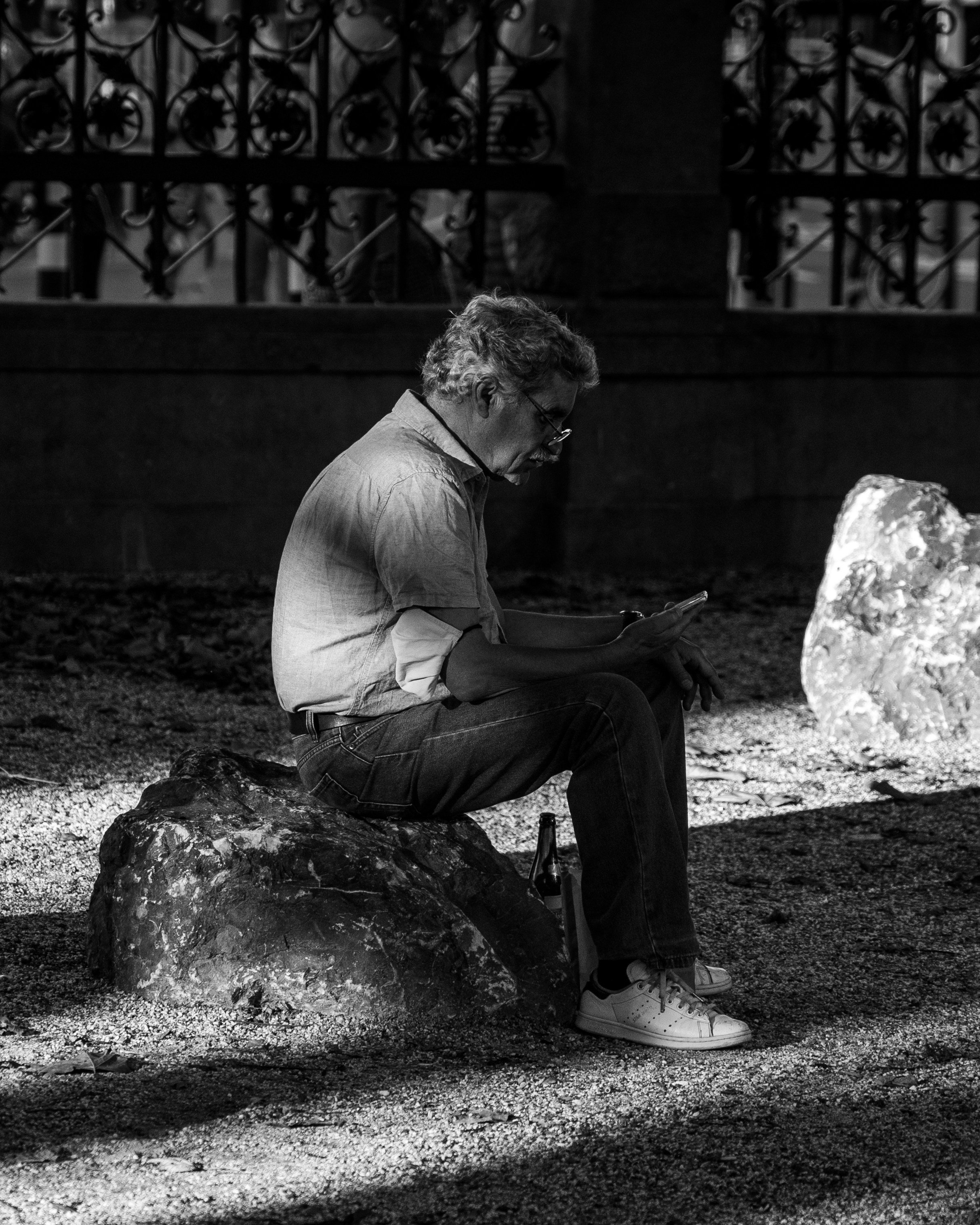Elderly man seated on a rock, engrossed in his phone, illuminated by soft sunlight amidst a shadowy park setting.