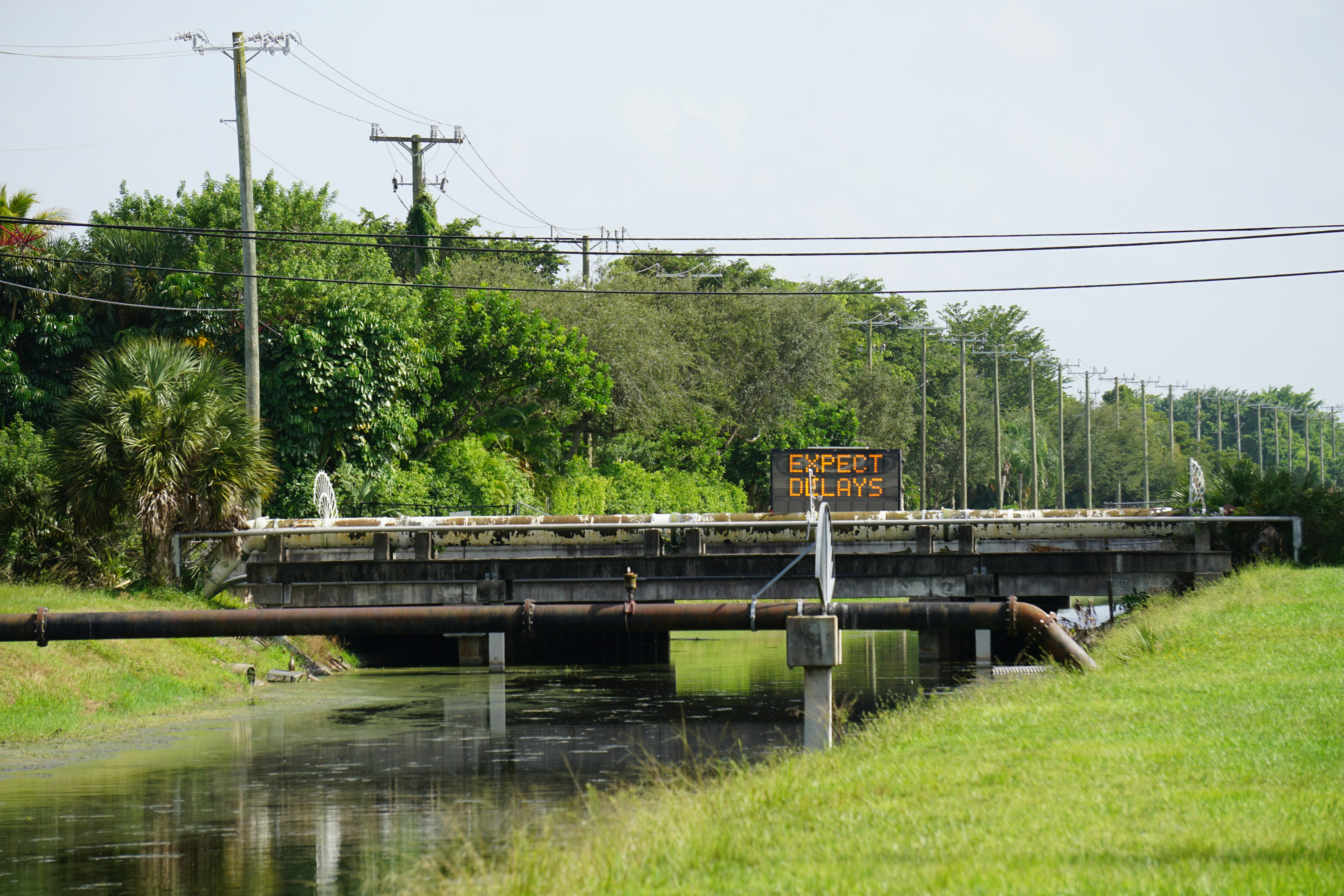 A bridge spans a canal, with a digital sign displaying 'EXPECT DELAYS' amidst lush greenery and power lines.