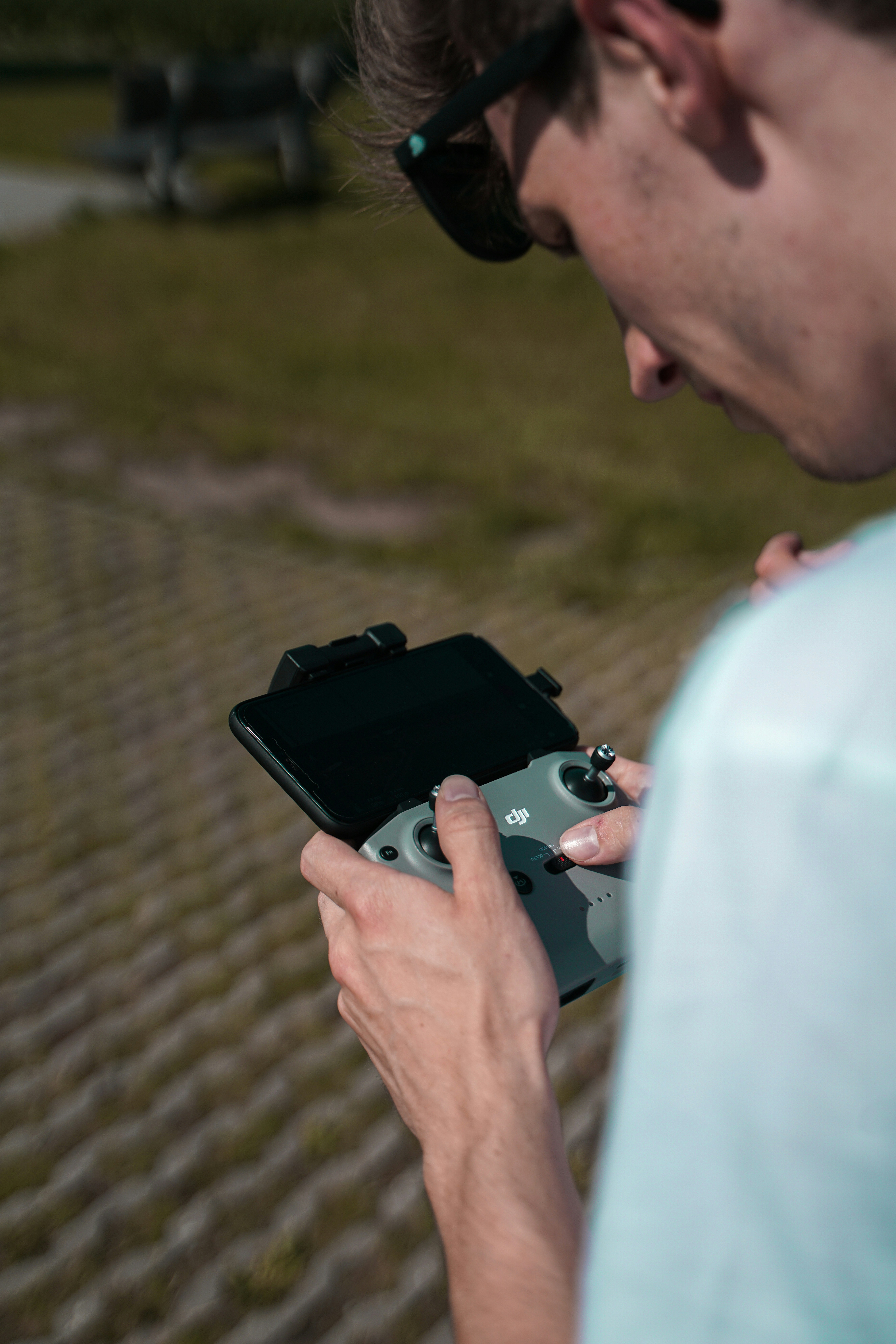 A person operating a drone controller with a smartphone attached, focused on the screen in an outdoor setting. The textured ground adds depth to the scene.
