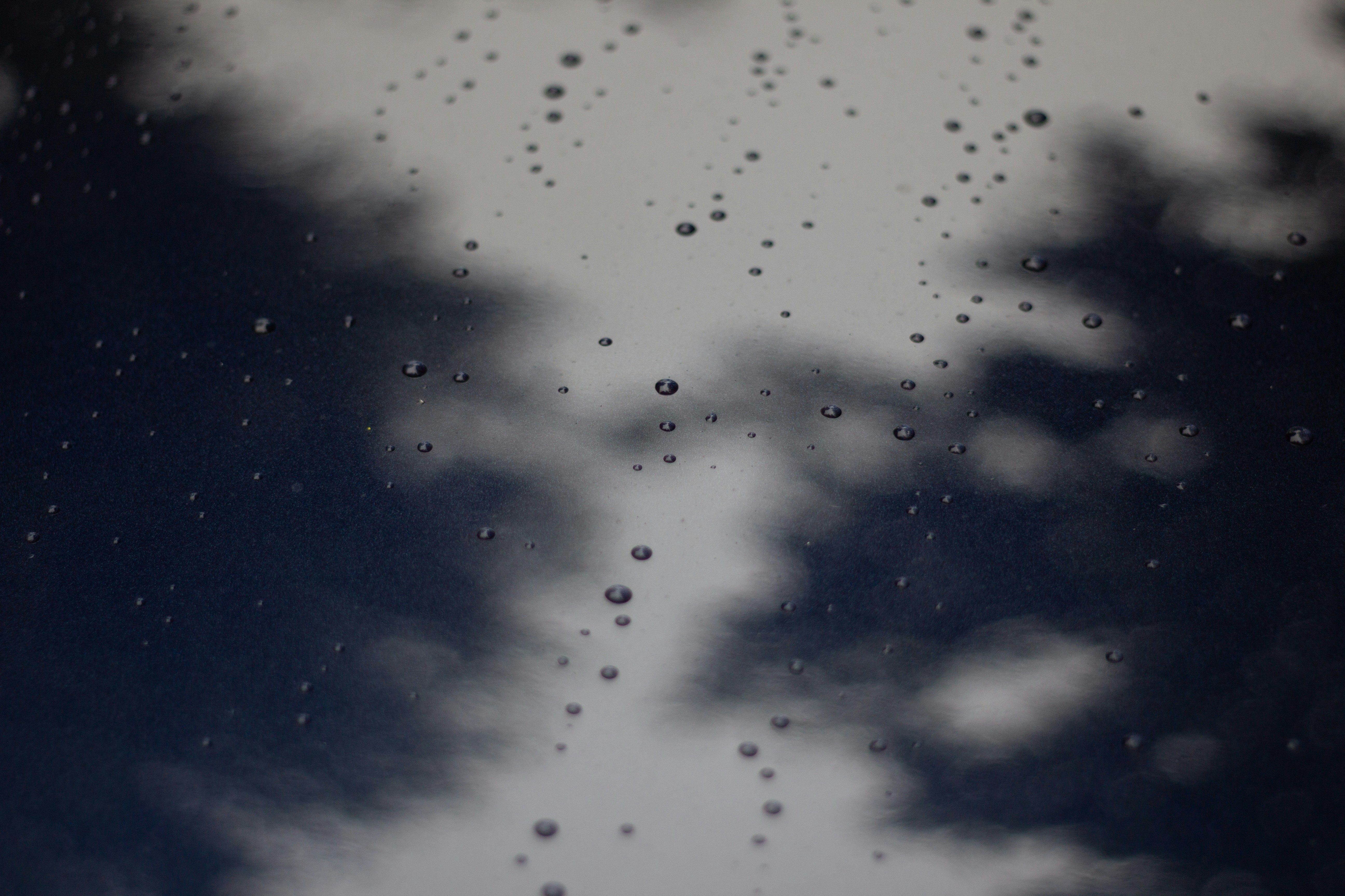 Raindrops cluster on a dark surface, creating a mesmerizing interplay of reflections and shadows. The scene captures the essence of a rainy day.
