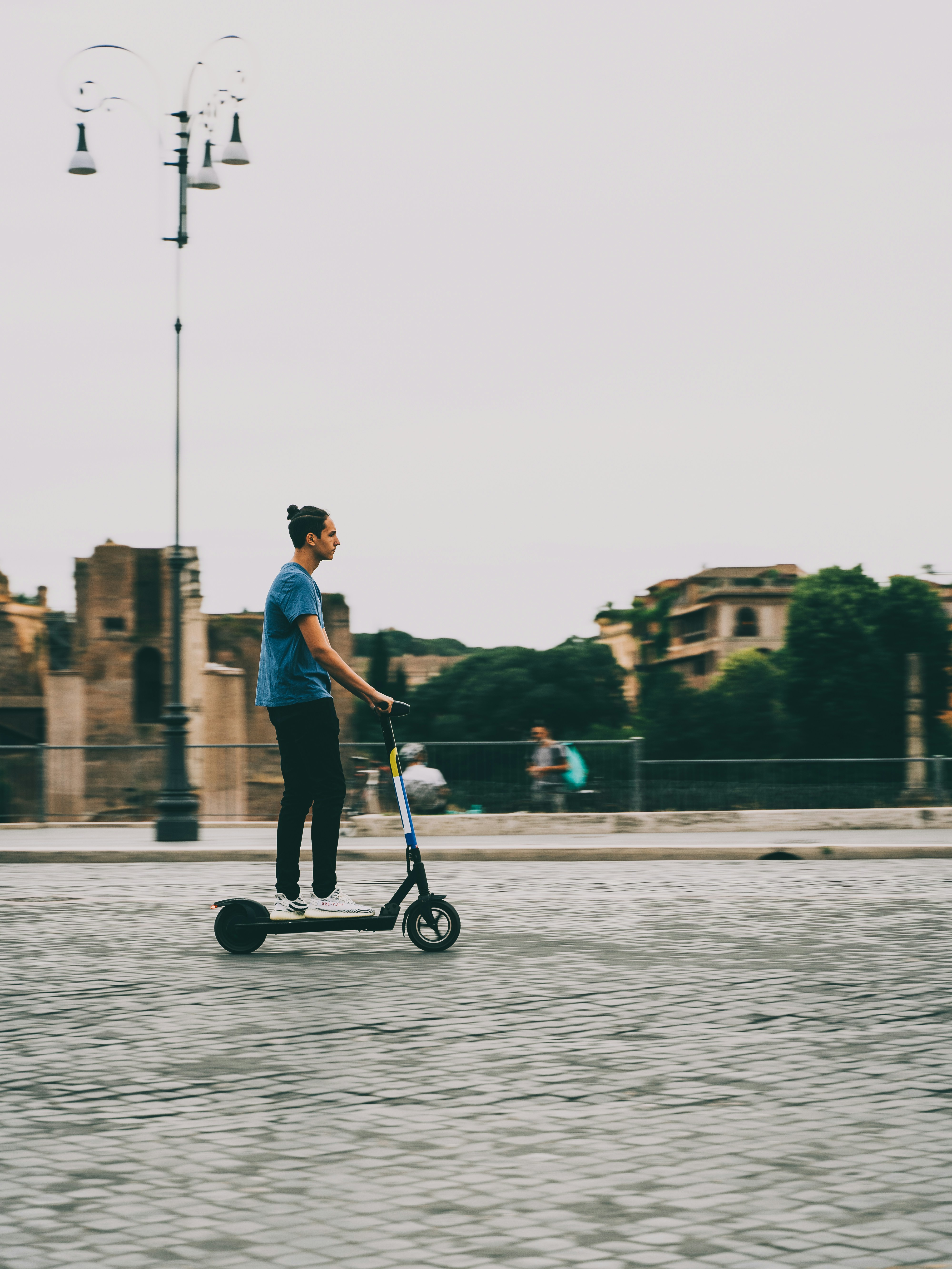 Young man riding an electric scooter past historic architecture in a bustling urban setting.