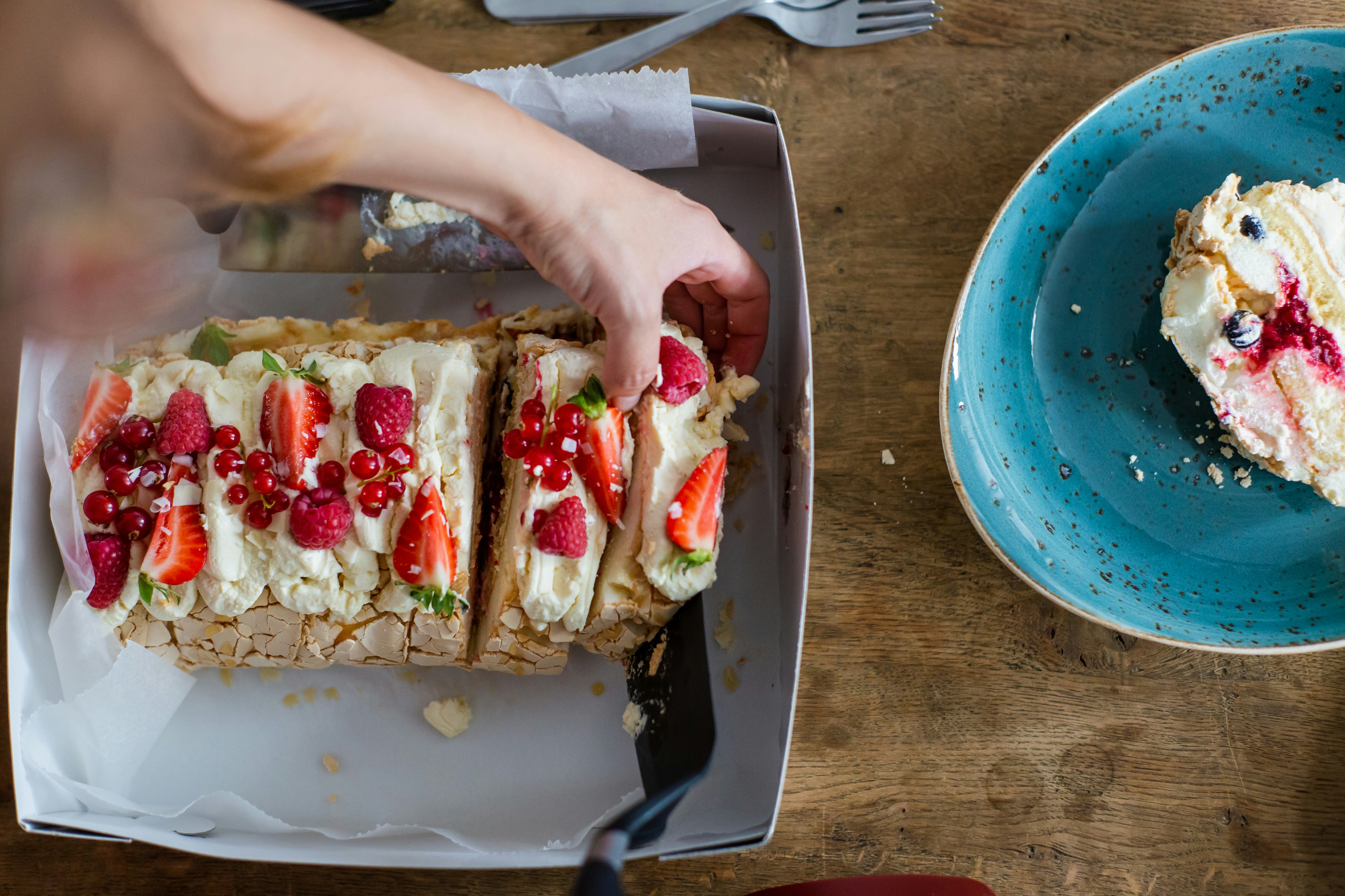 Hand reaching for a slice of cake adorned with fresh strawberries and pomegranate seeds, next to a turquoise plate on a wooden table.