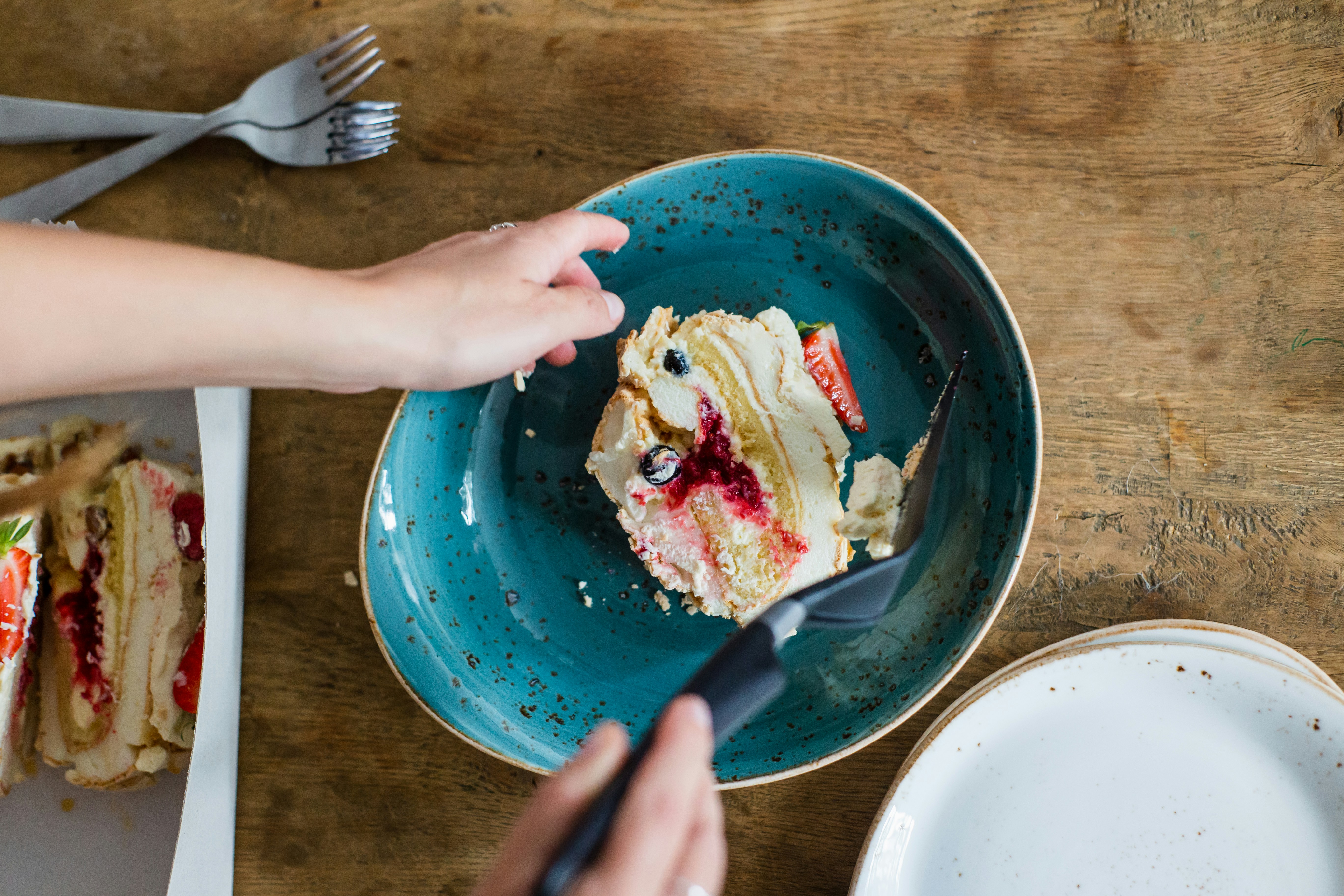 Hands serving a slice of berry-topped cake onto a turquoise plate on a wooden table.