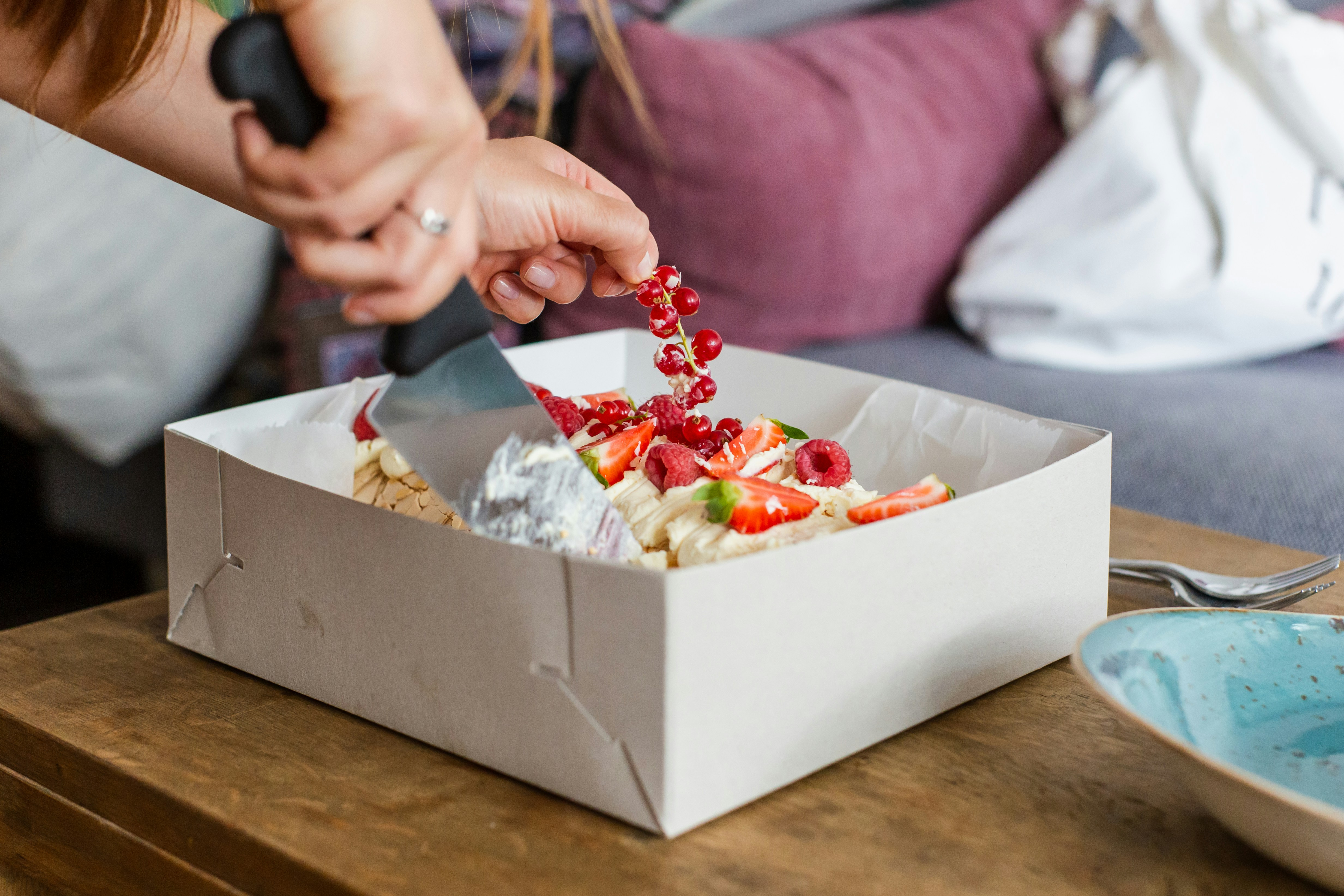 Hands slicing pomegranate seeds over a decorative dessert in a white box on a wooden table.