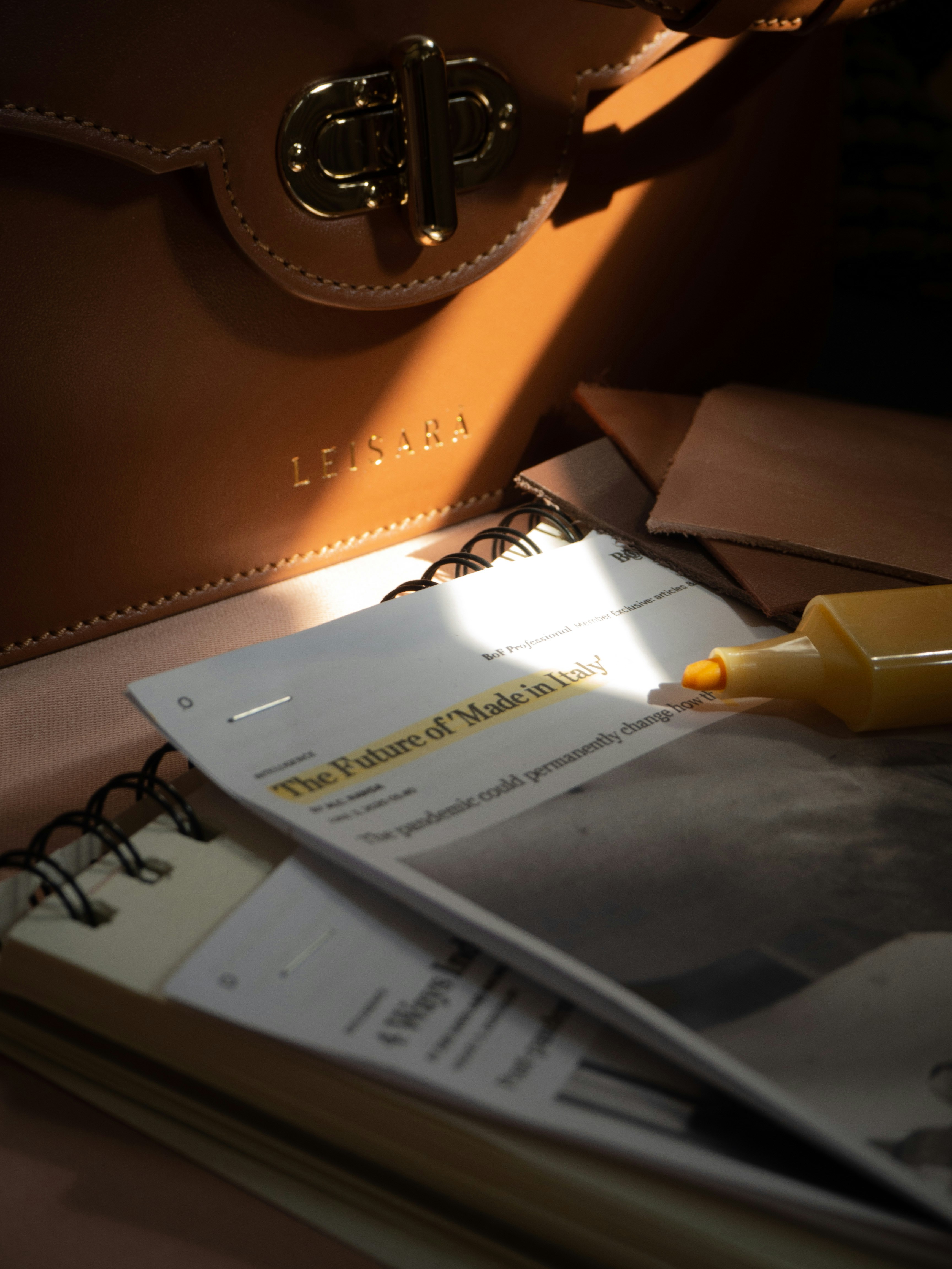 A stylish leather handbag rests beside an open notebook and a highlighter, with sunlight casting soft shadows across the scene.