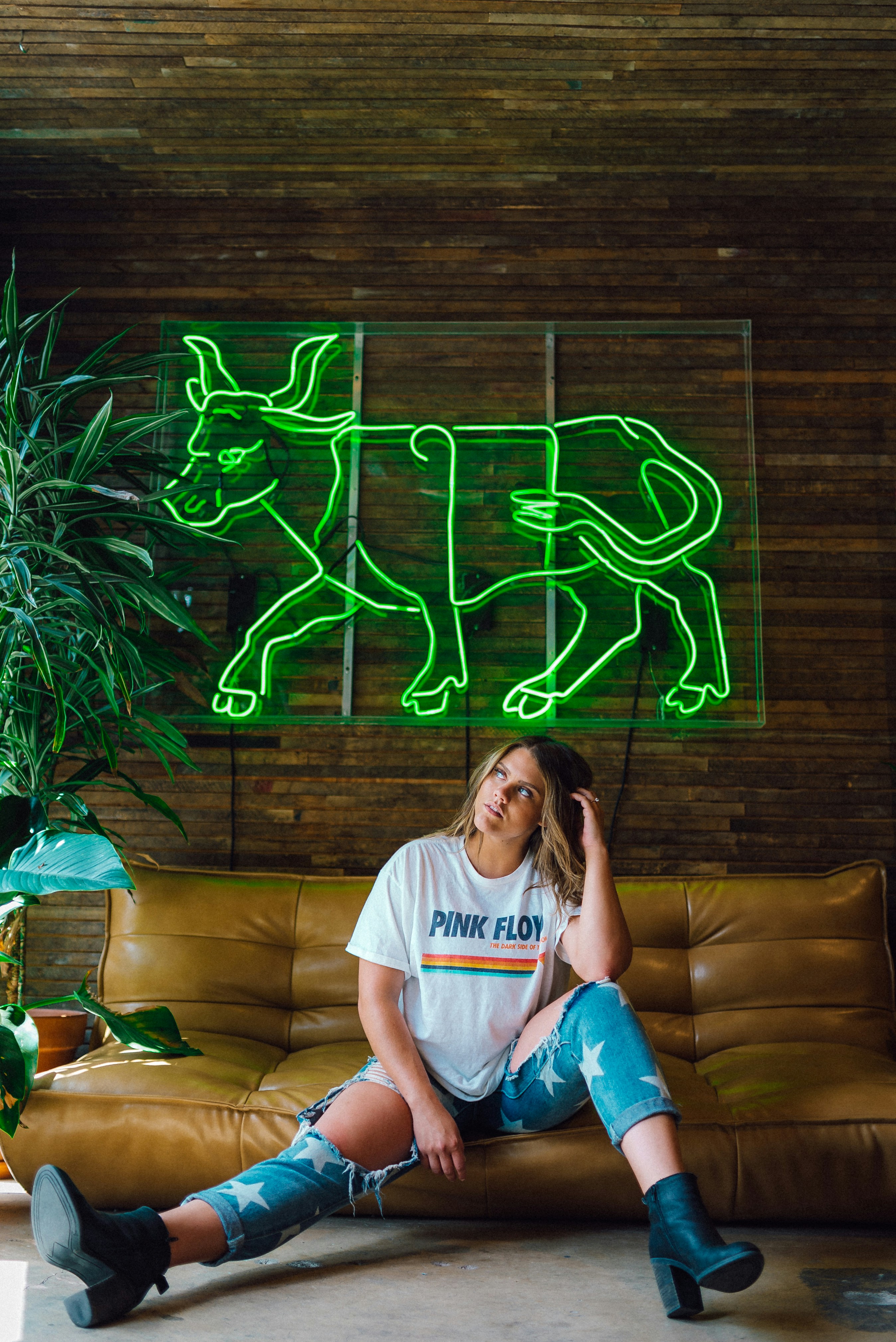 A young woman in a graphic tee poses thoughtfully on a couch, illuminated by a vibrant neon cow sign against a rustic wooden backdrop.
