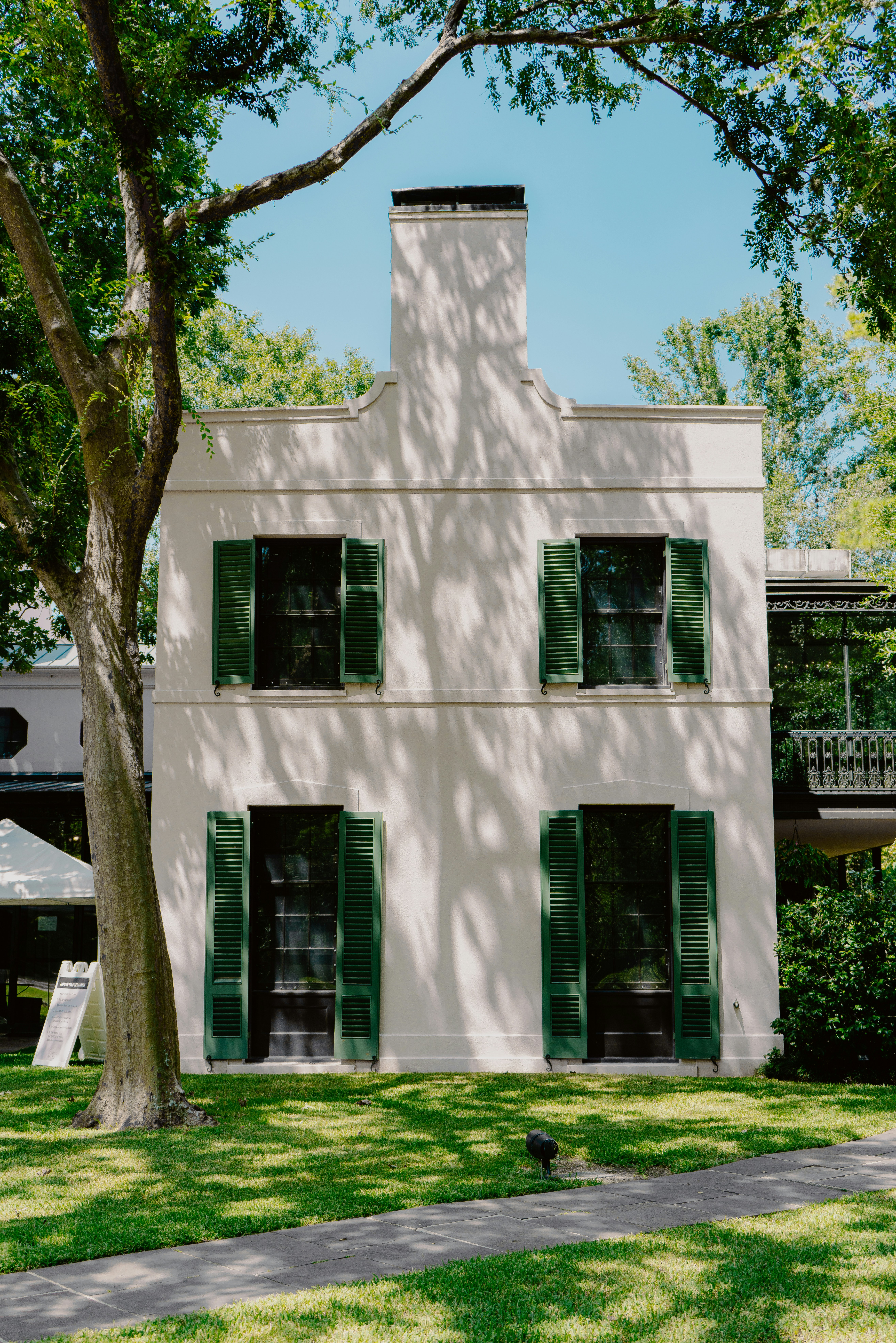 White building with green shutters framed by lush trees casting dappled shadows.